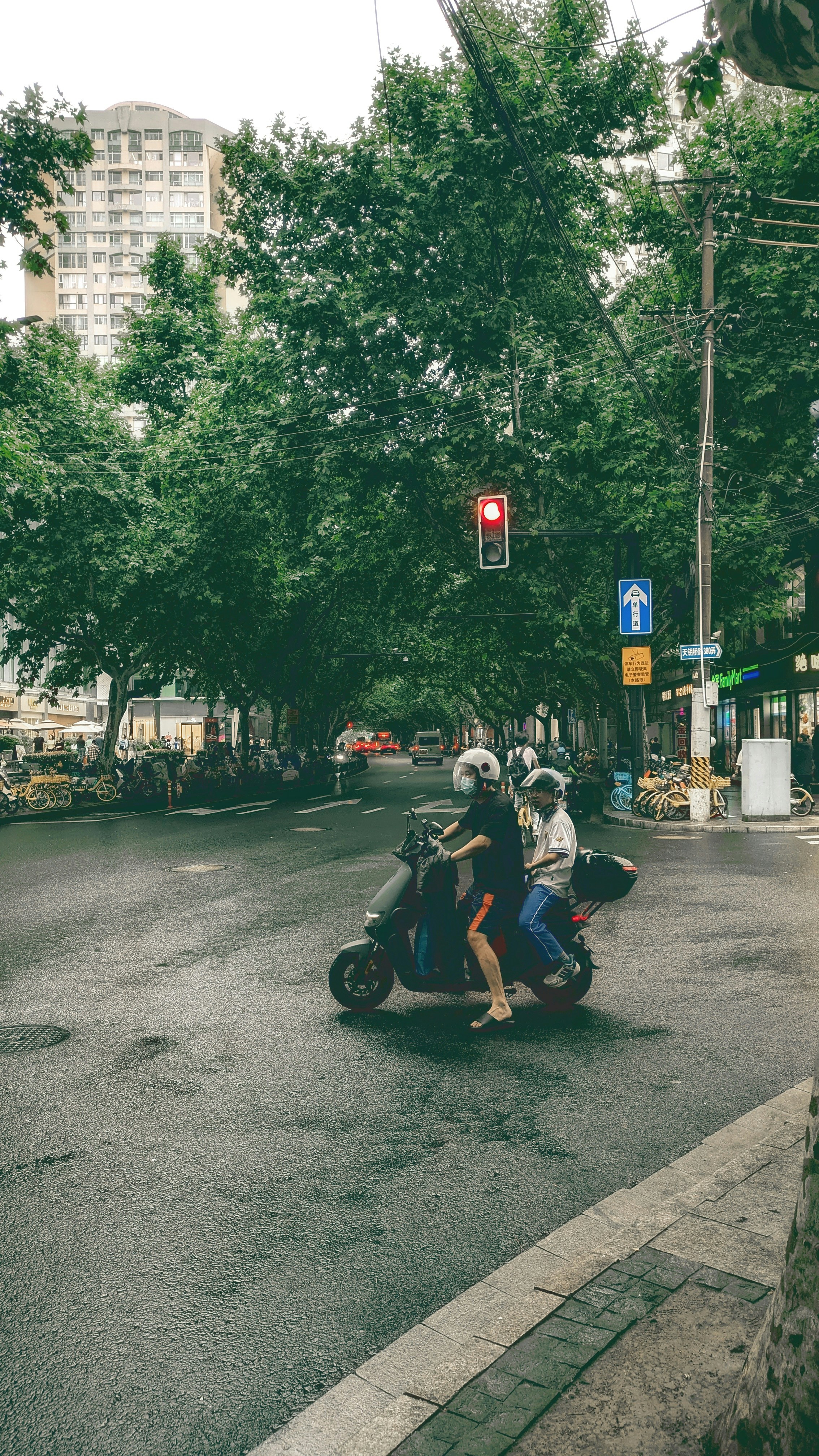 Two riders on a scooter pause at a busy urban intersection, framed by lush trees and a red traffic light in the background. A street scene with storefronts and pedestrians adds context to a late-afternoon commute.