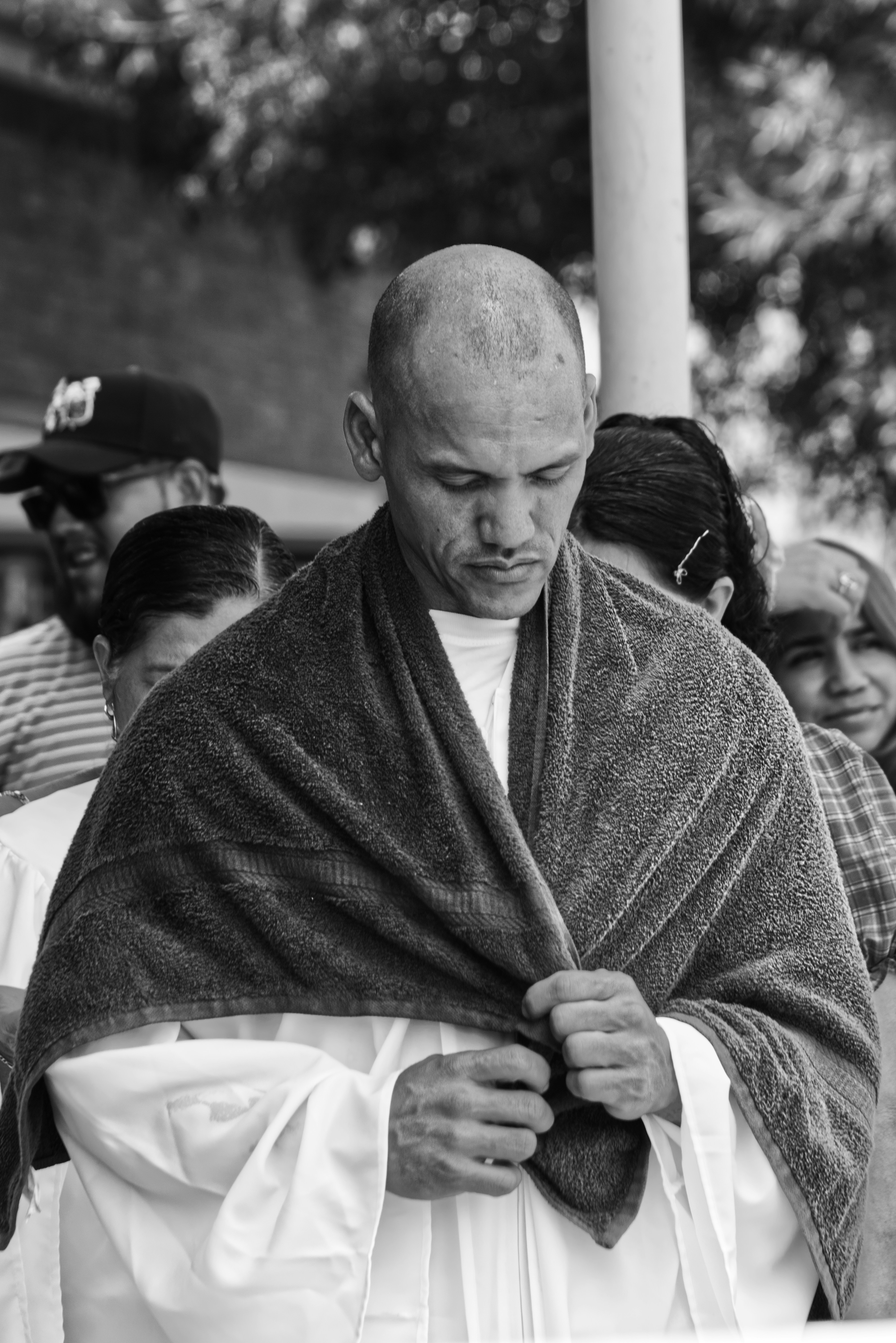 Monk in white robes wrapped in a towel stands with head bowed, surrounded by onlookers in a grayscale crowd.