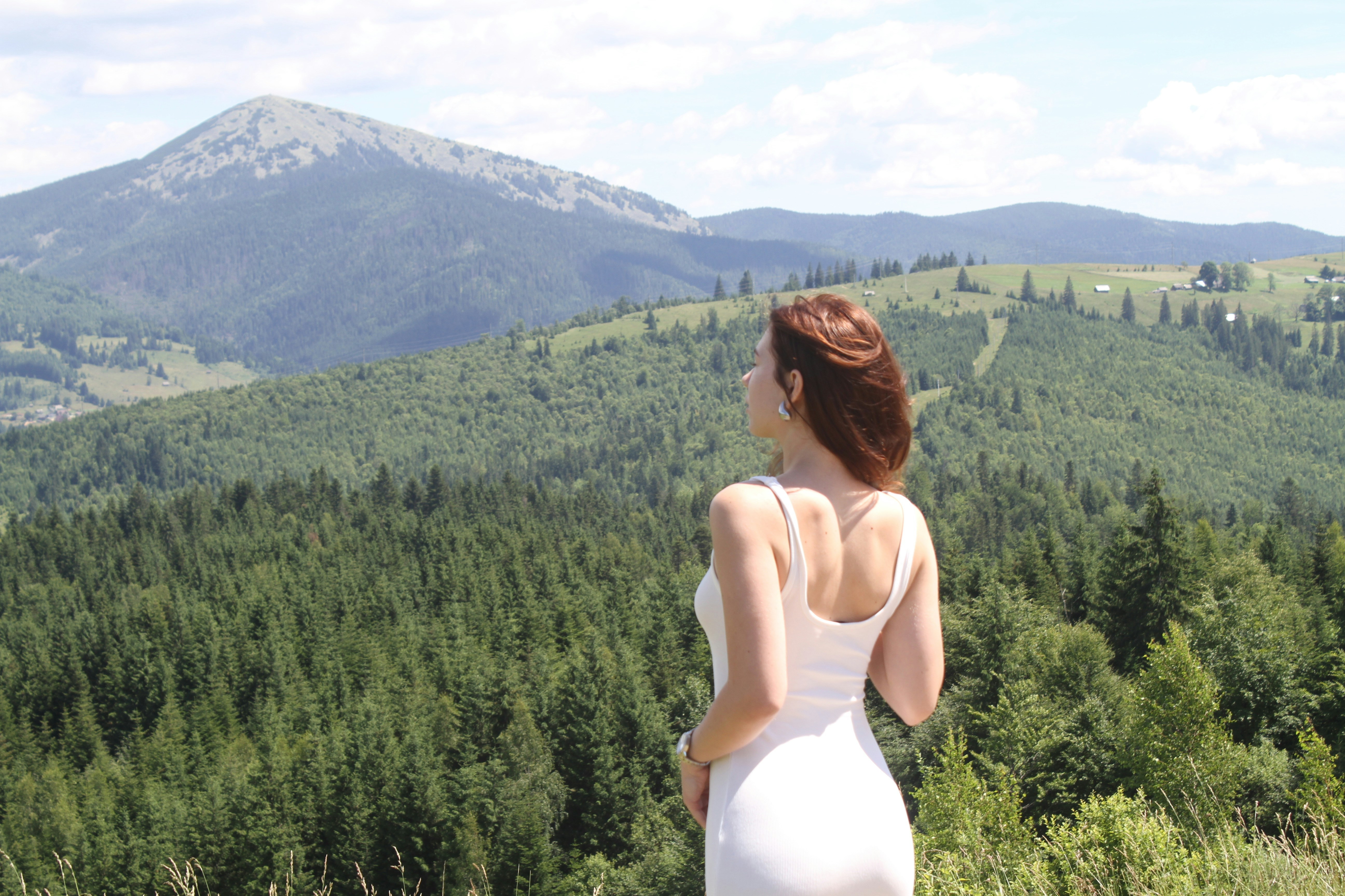 A woman in a white dress stands with her back to the camera, gazing toward a forested valley and distant mountains, conveying a serene hillside landscape.