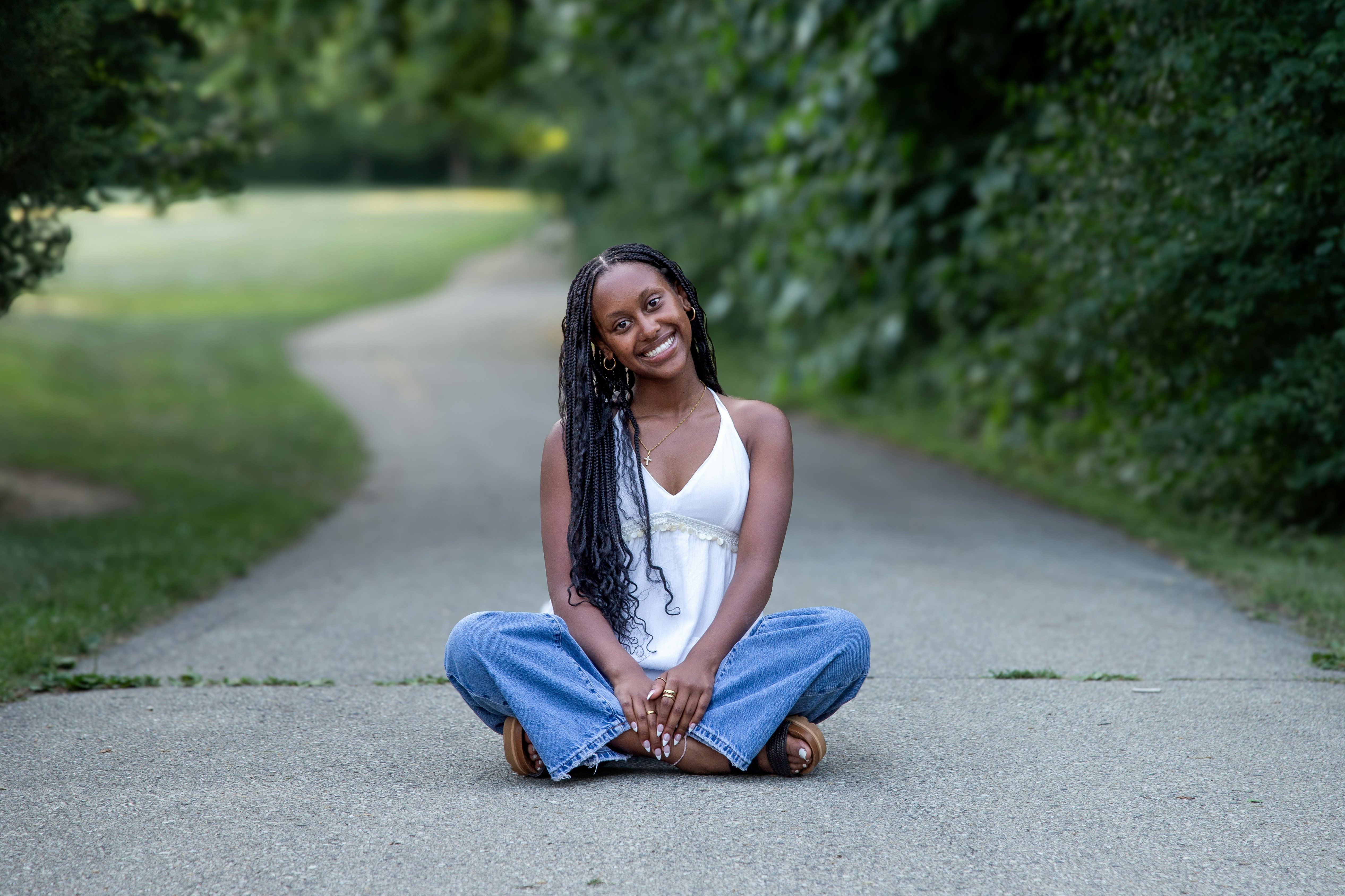Girl seated cross-legged on a sunlit park road, smiling toward the camera