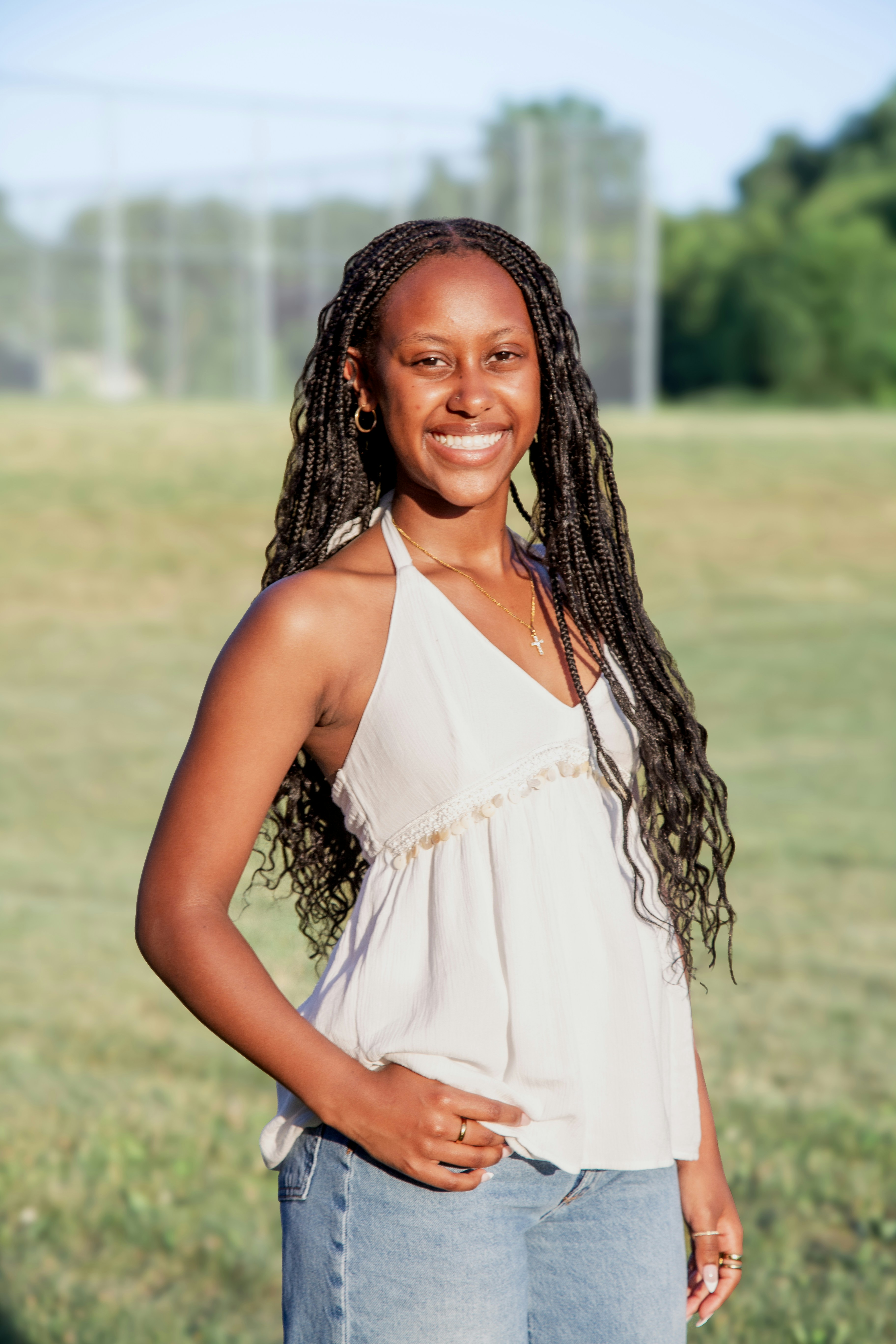 A smiling woman stands outdoors on a sunlit field, wearing a white halter top and denim jeans. The image captures a warm, candid portrait with a softly blurred background, emphasizing the subject's cheerful presence.