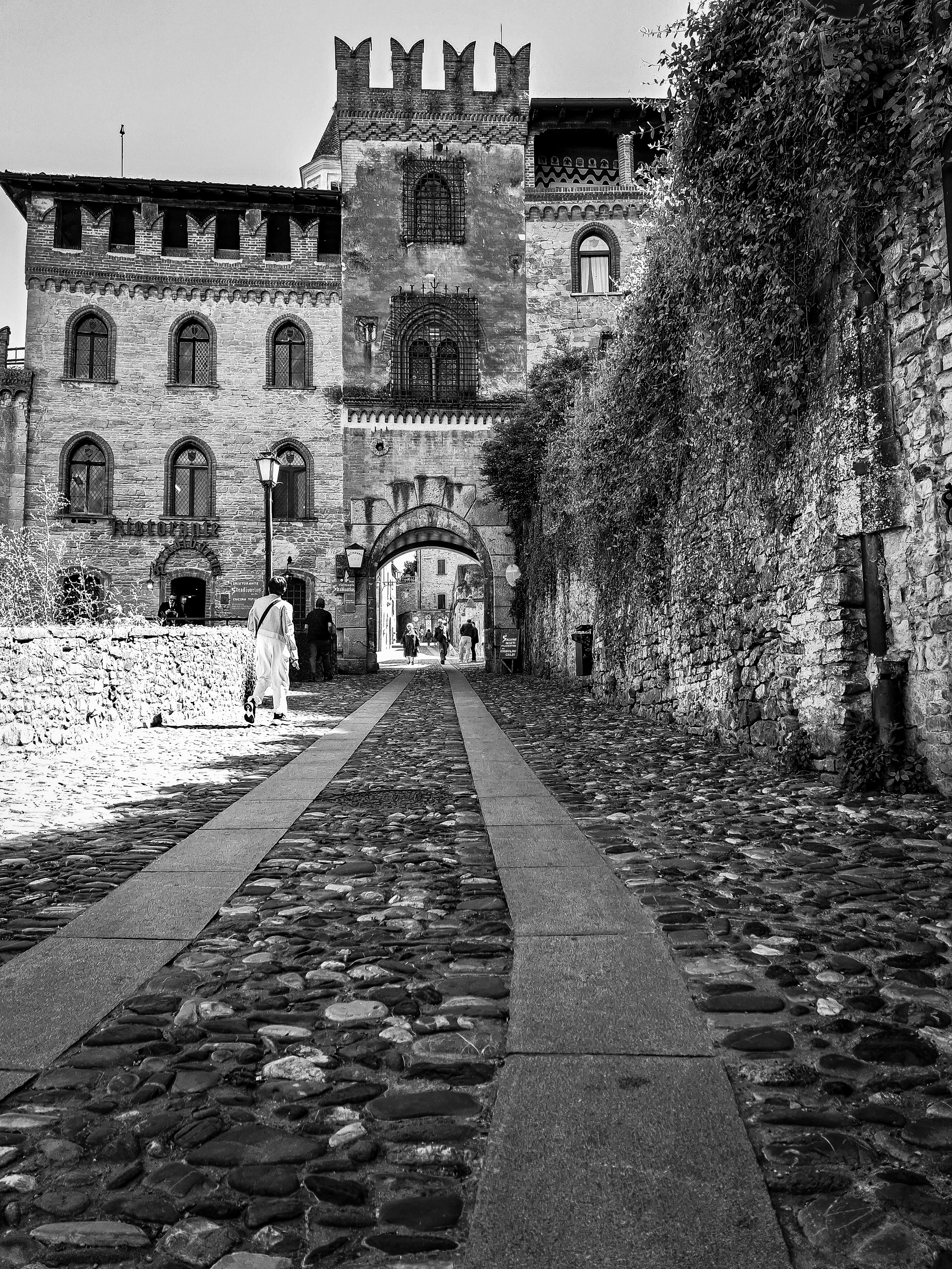 Cobblestone street leads through a historic stone archway toward a distant courtyard, framed by aged fortress walls and ivy-clad ramparts.