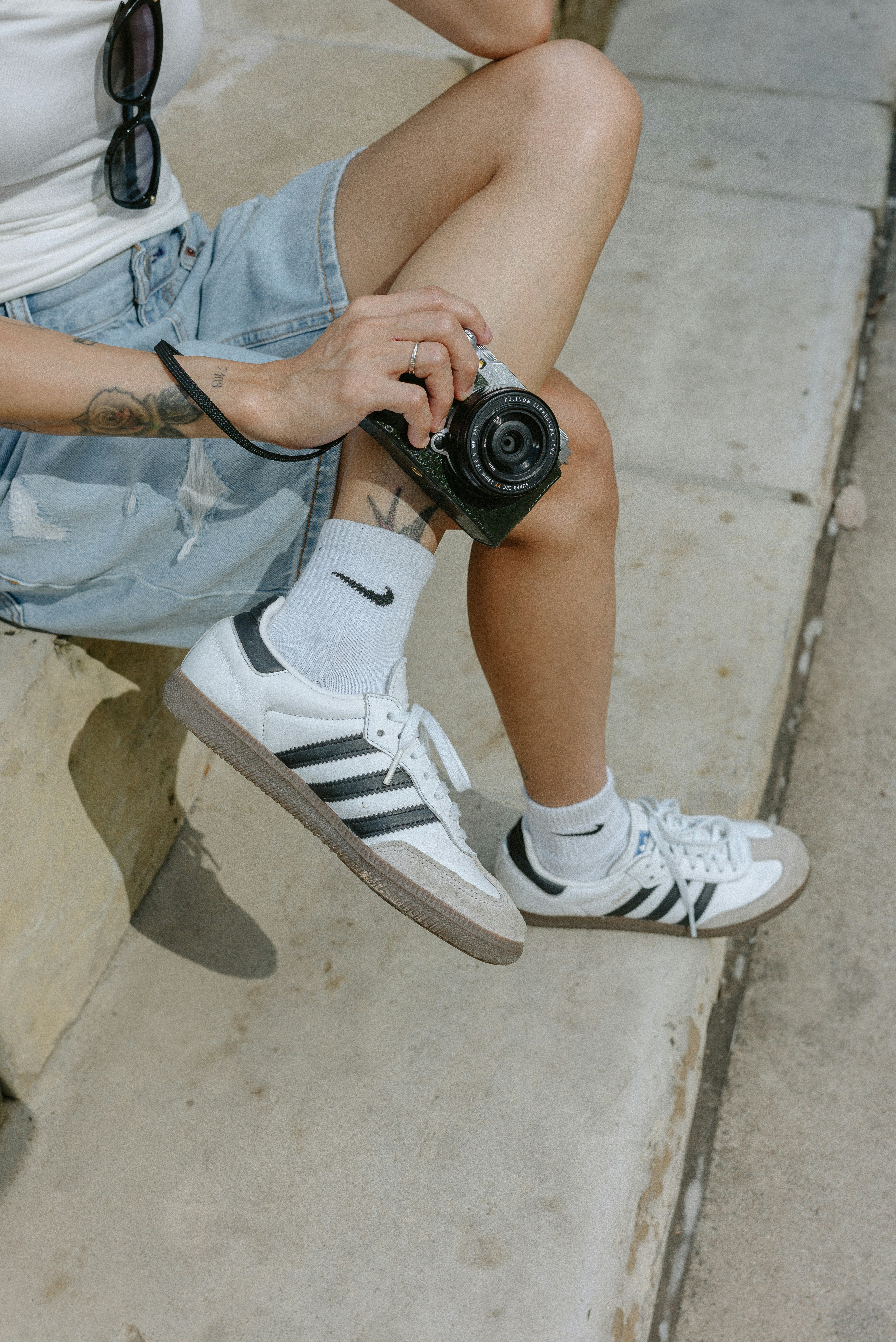 Casual portrait of a person seated on a concrete ledge, holding a compact camera; sneakers, denim shorts, and sunglasses add sporty details to the scene. The composition emphasizes the camera as the focal point and a relaxed, candid vibe.