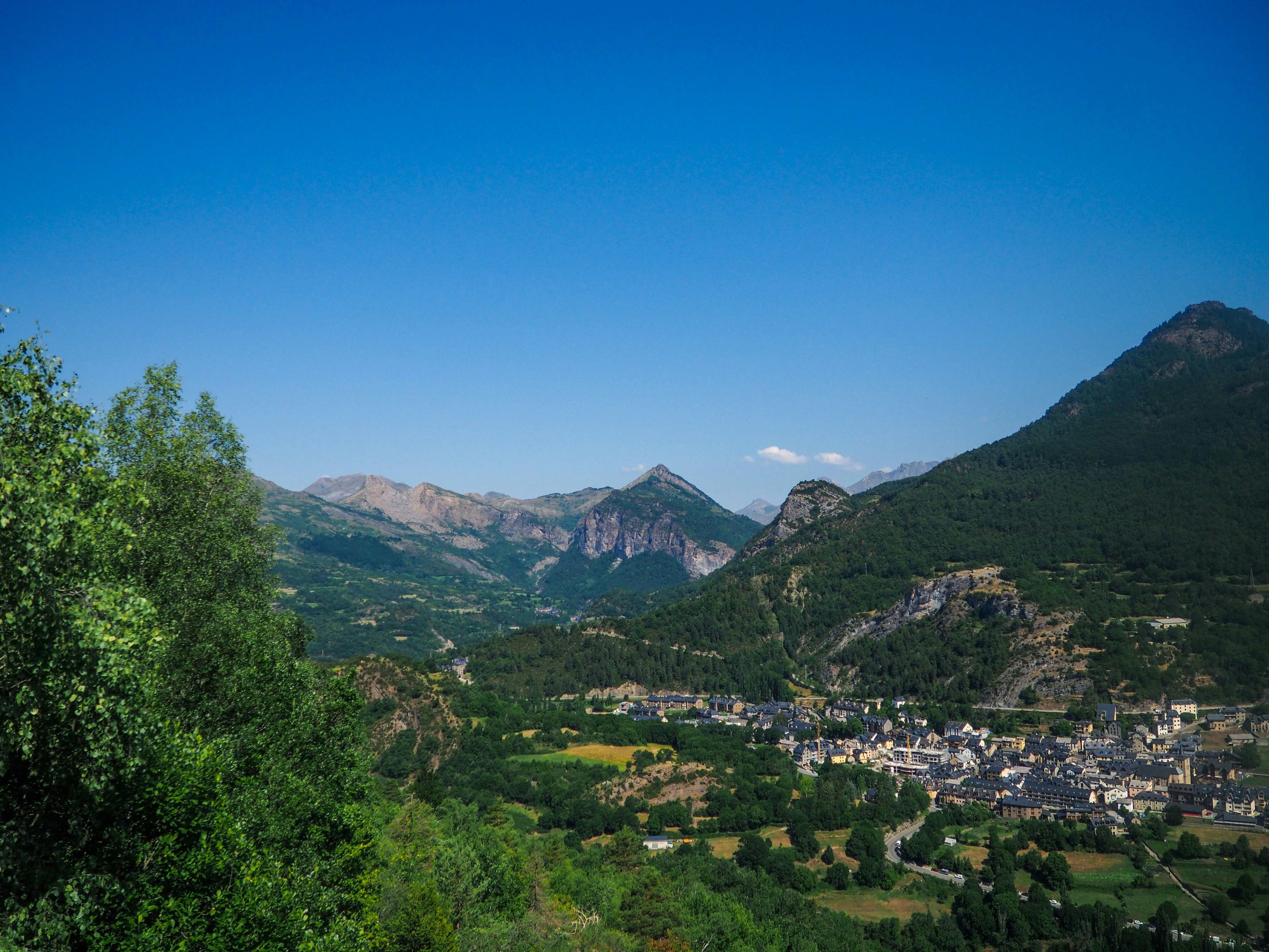 Expanse of a sunlit alpine valley with a clustered village at the foothills and surrounding green terraces under a clear blue sky.