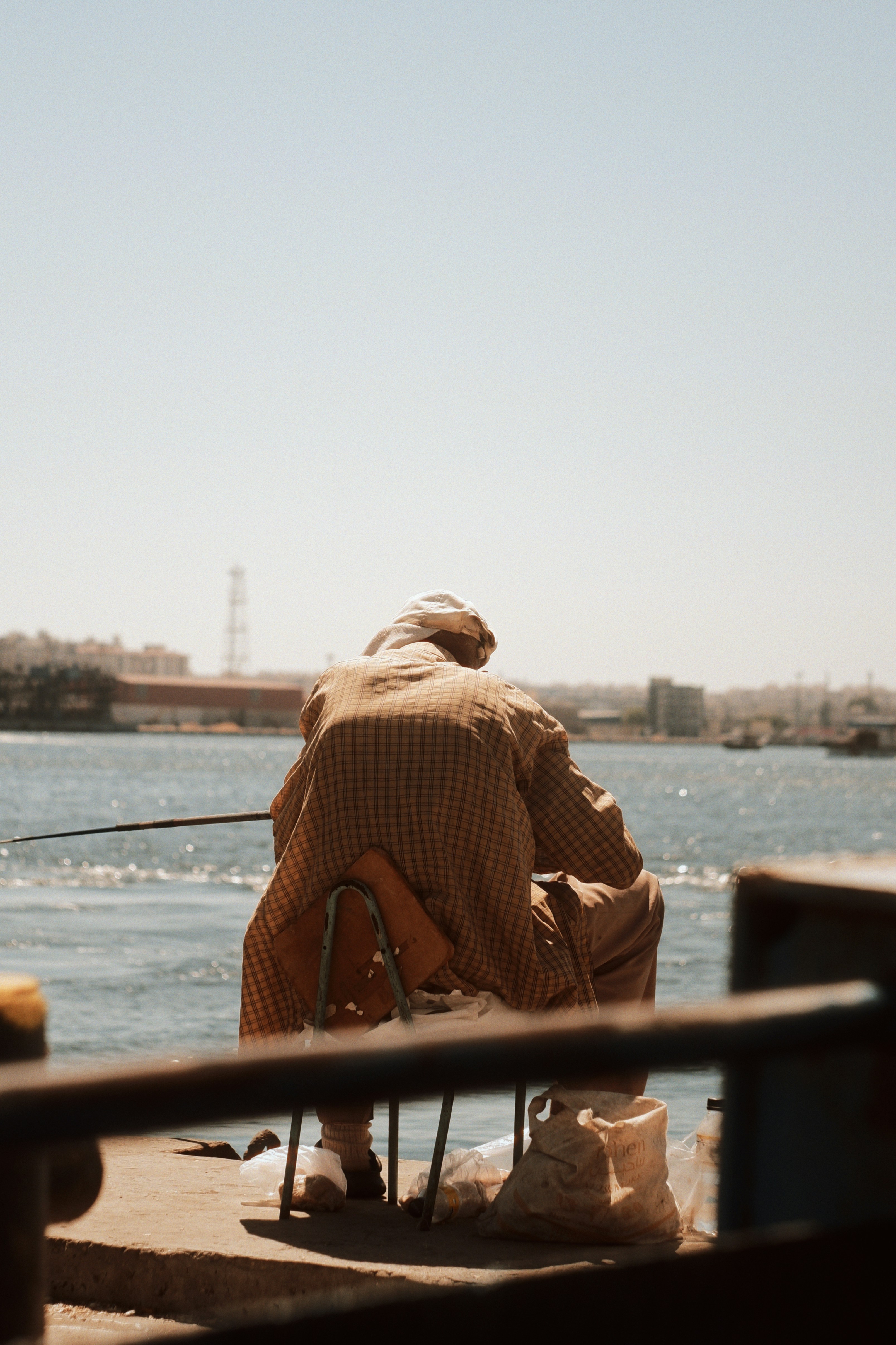 A solitary fisherman in a checked cloak sits on a chair at the edge of the wharf, casting a line toward sunlit water with distant buildings on the horizon.