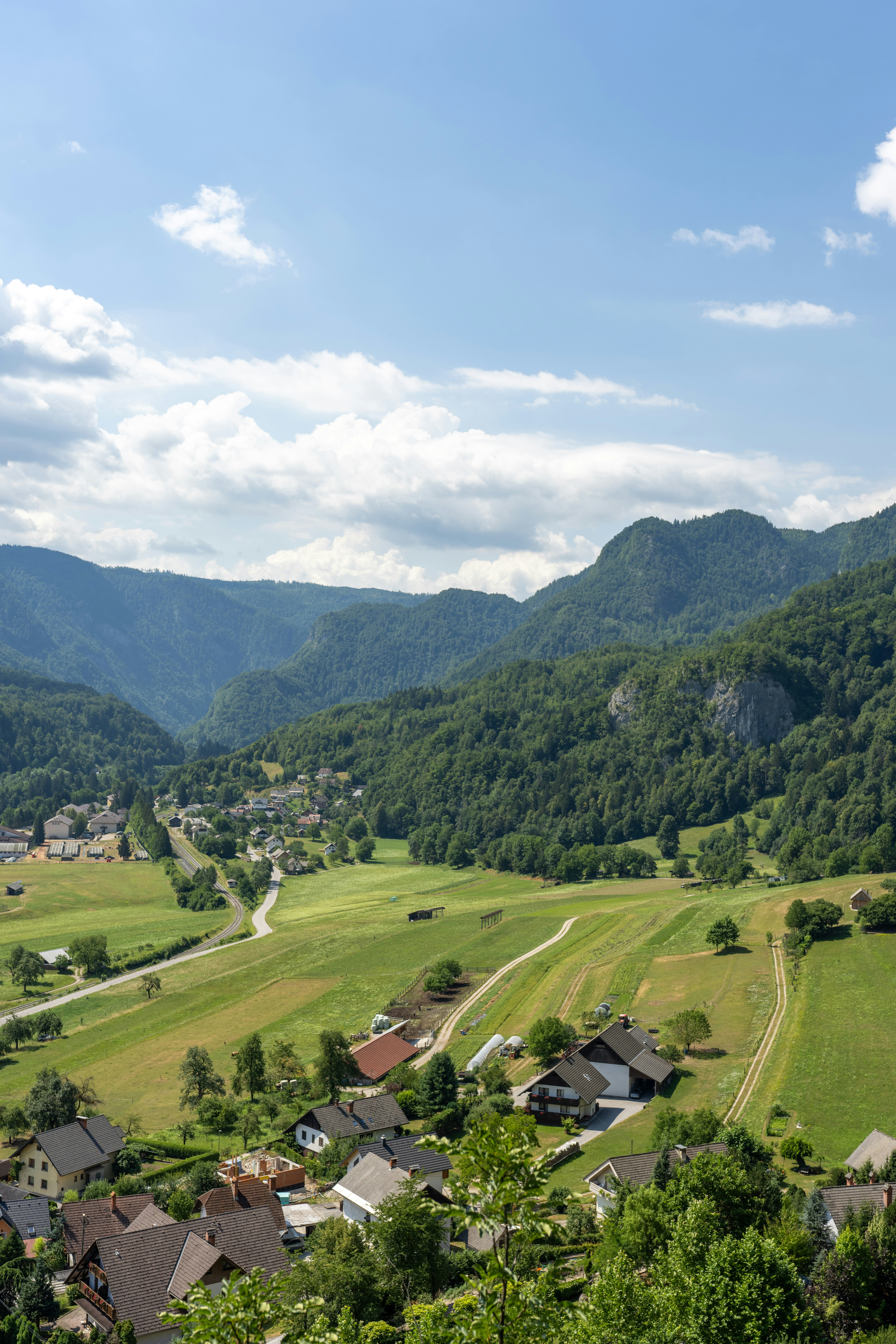 Sunlit valley with a cluster of houses and winding roads framed by forested hills and distant mountains.