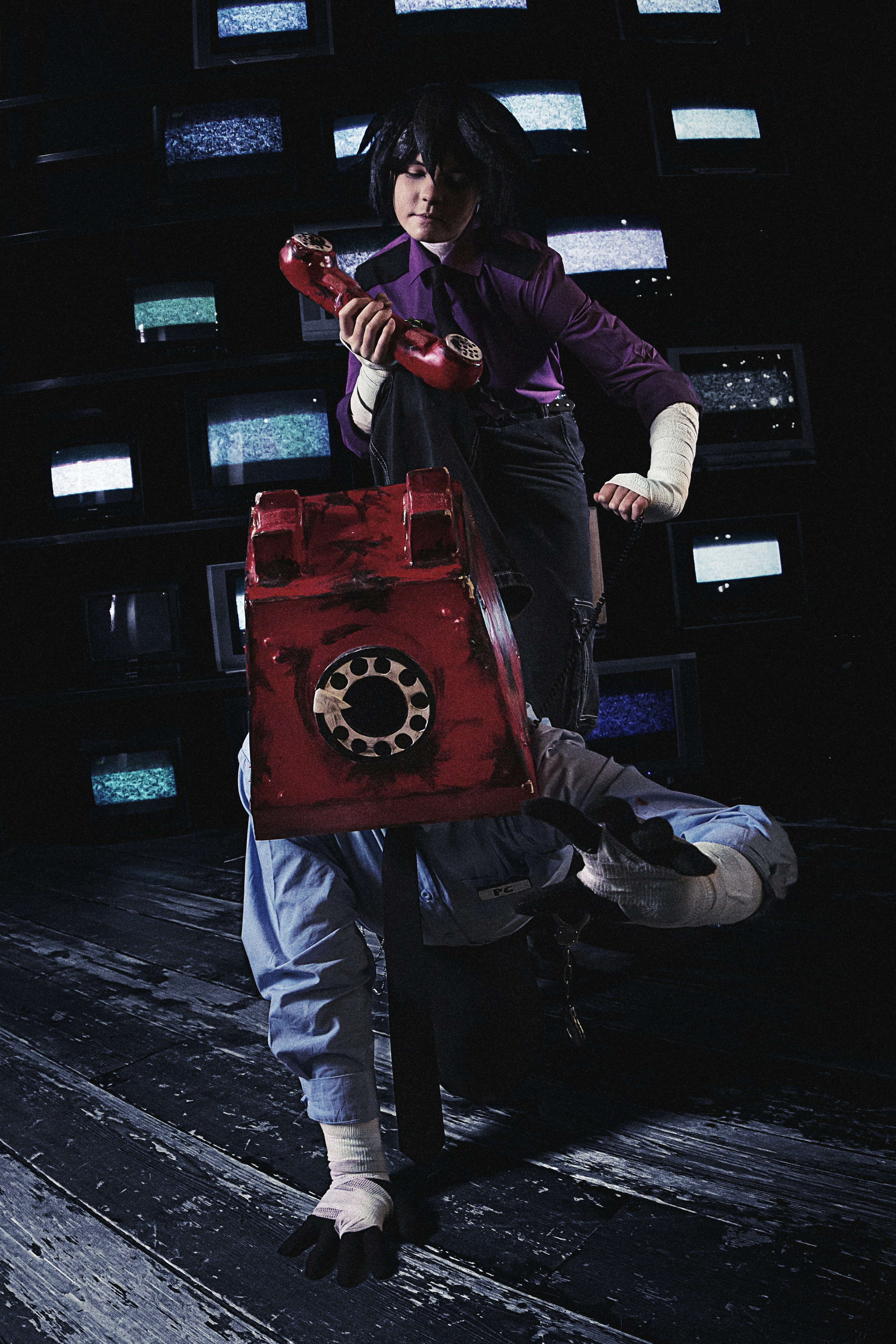 A person in a purple shirt grips a red vintage telephone while kneeling among stacked TVs in a dark room; dramatic lighting highlights the subject and props to create a moody, surreal scene.