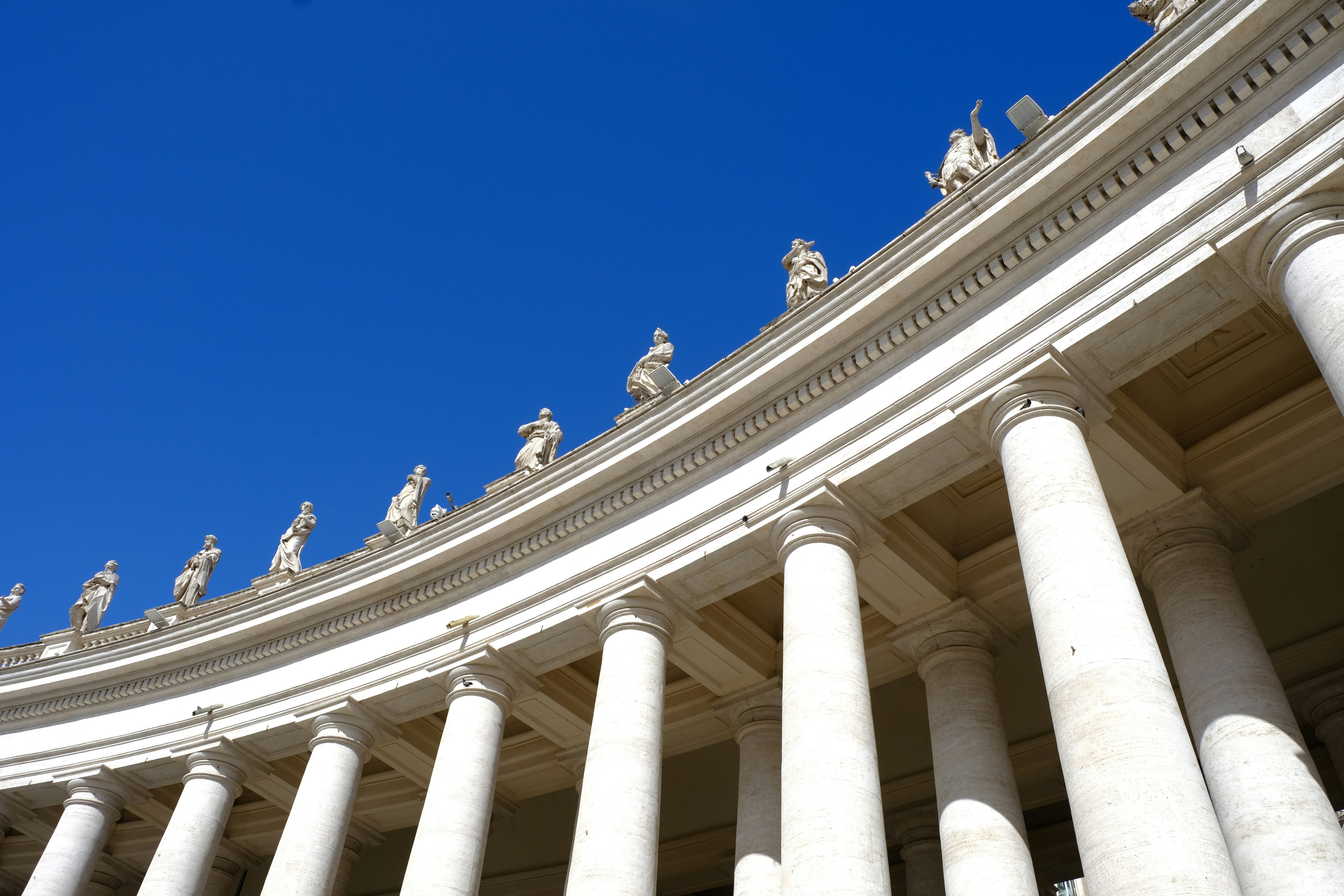 Curved row of statued pedestals atop grand white columns against a bright blue sky; architectural detail and perspective highlight a monumental neoclassical facade.