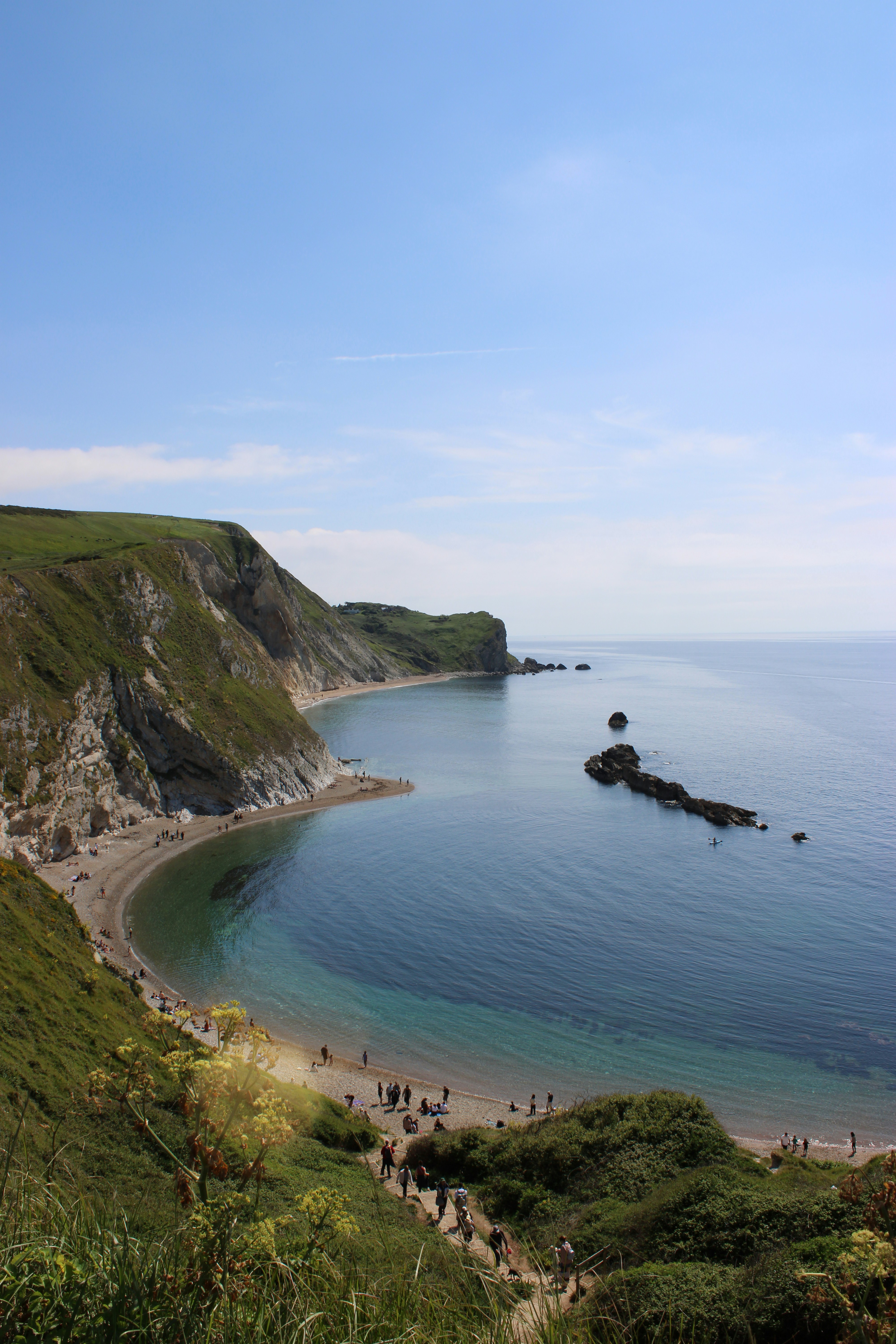 Sunlit coastal cove with sandy beach curved along emerald cliffs and a path of people descending to the shore amid clear blue water.