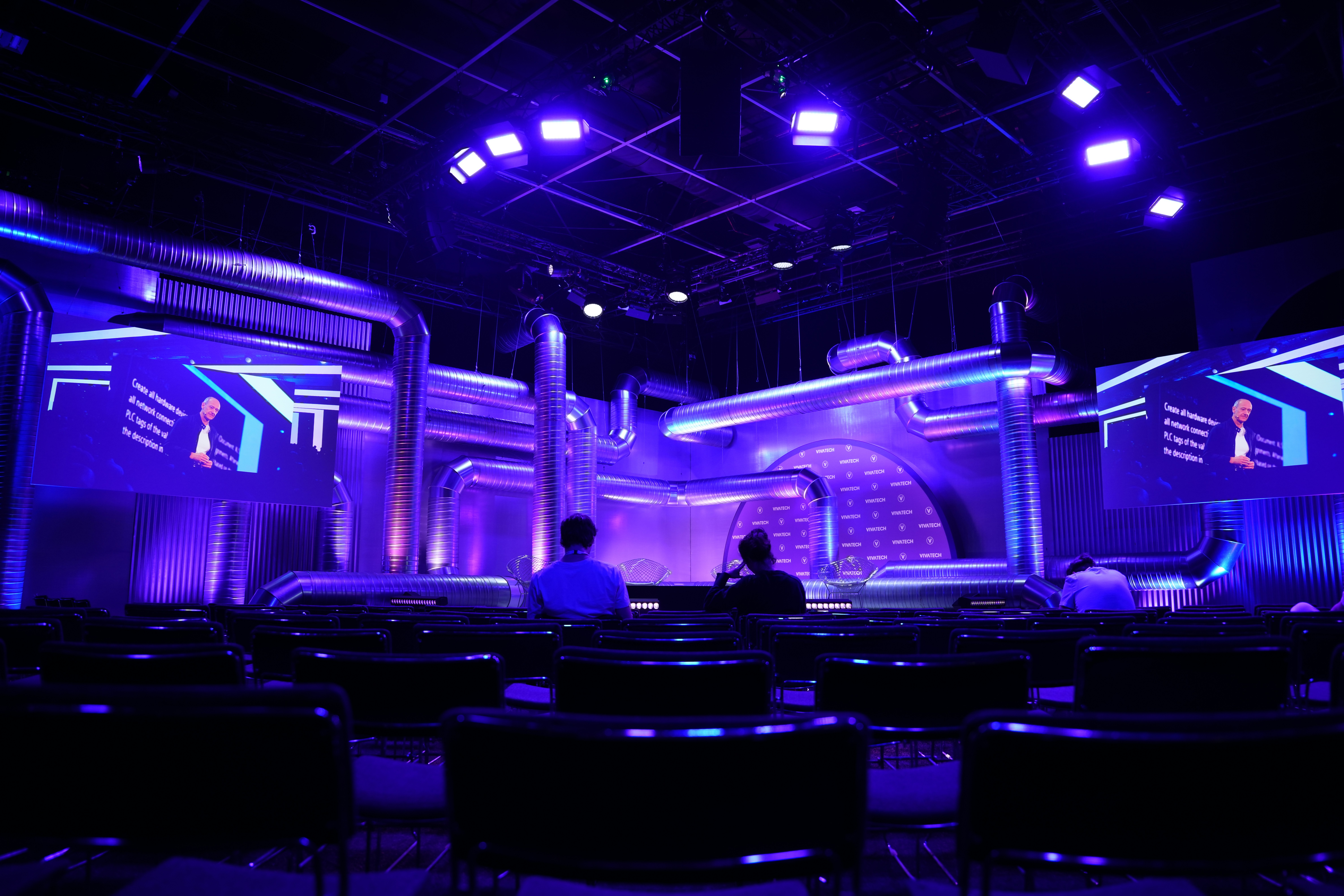 Dramatic, neon-lit backstage arena with two figures seated facing a glowing stage, framed by futuristic ducts and screens. A performance space set for a show, with moody blue lighting and rows of empty seats.