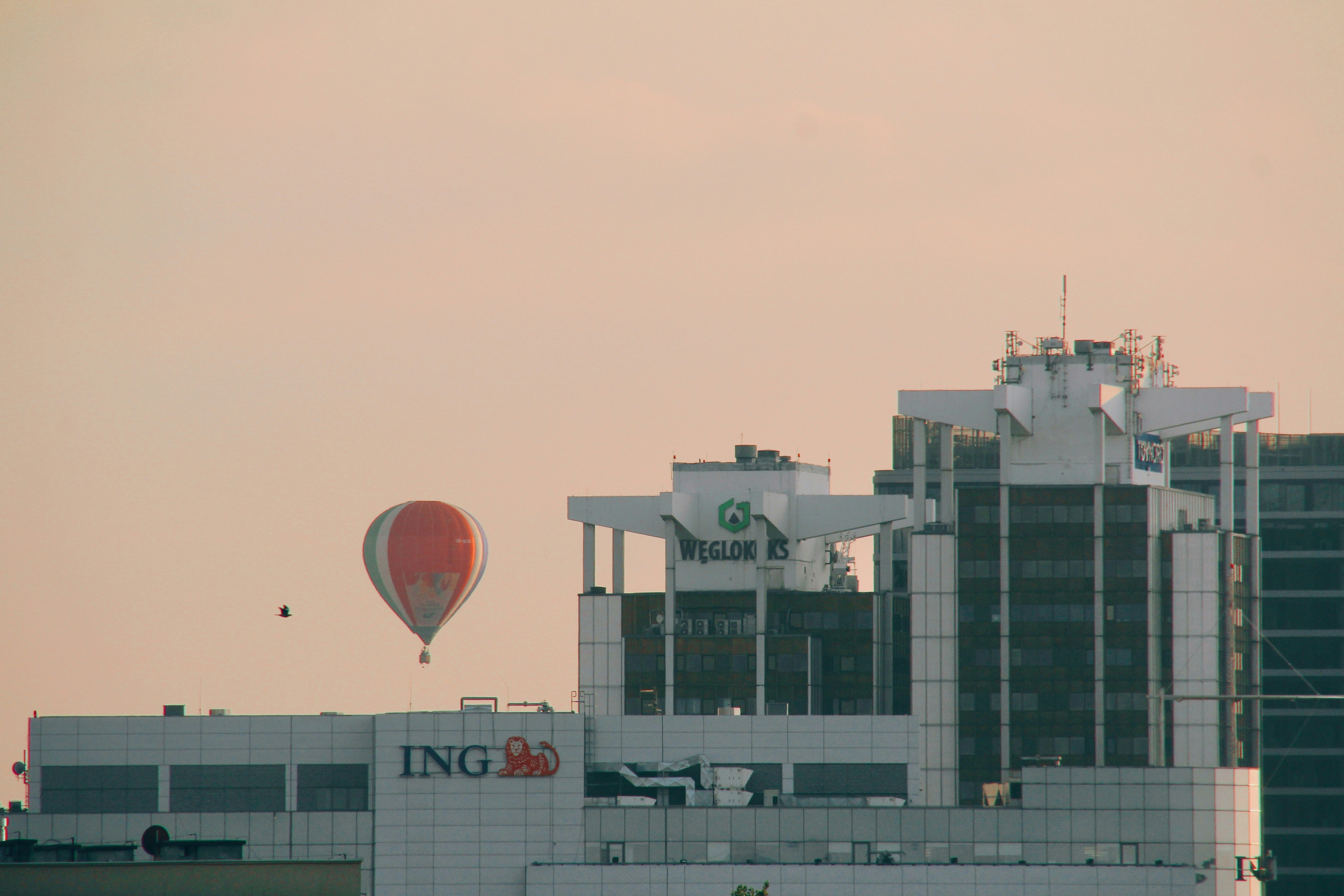 Balloon drifts above a modern corporate complex at dusk, with signage such as ING and WELOK visible on the rooftops.