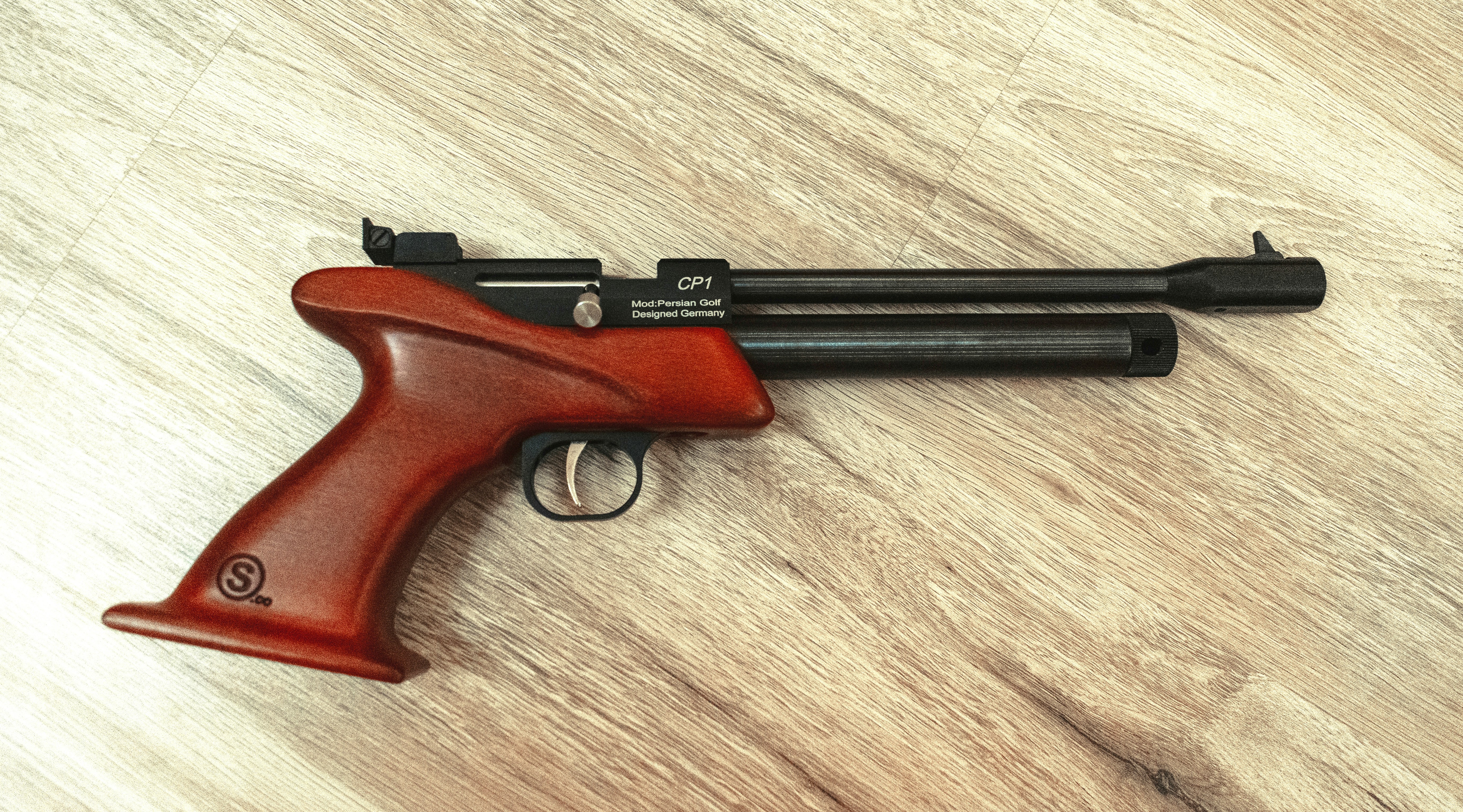 Compact air pistol with a wooden grip sits on a pale wooden floor, highlighting its form and metallic barrel. A simple studio-like scene focusing on the tool's silhouette.