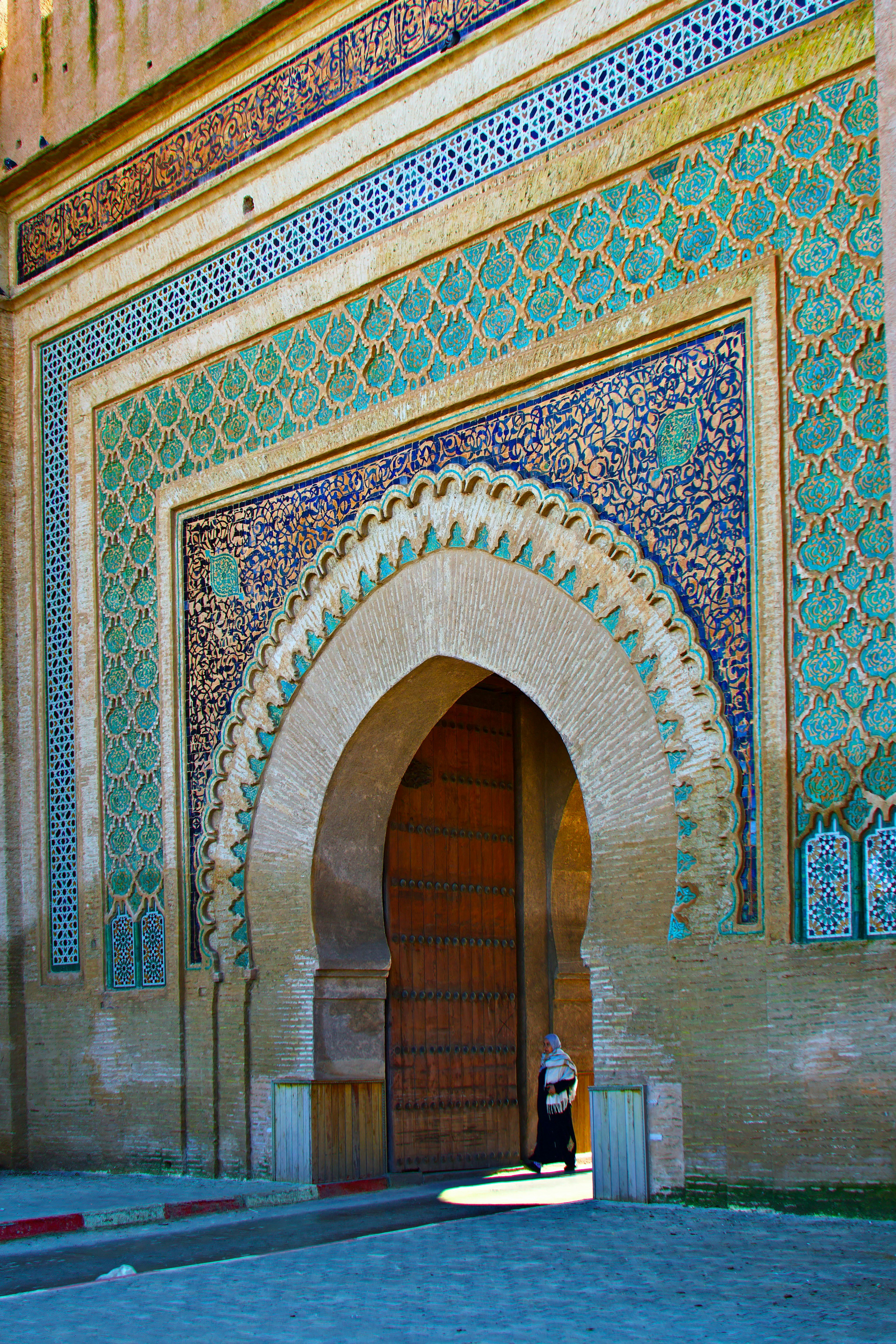  Ornate tiled archway with a wooden door forms the focal frame; a lone person stands at the doorway, casting a small shadow on the steps. The scene emphasizes architectural patterning and entryway scale.
