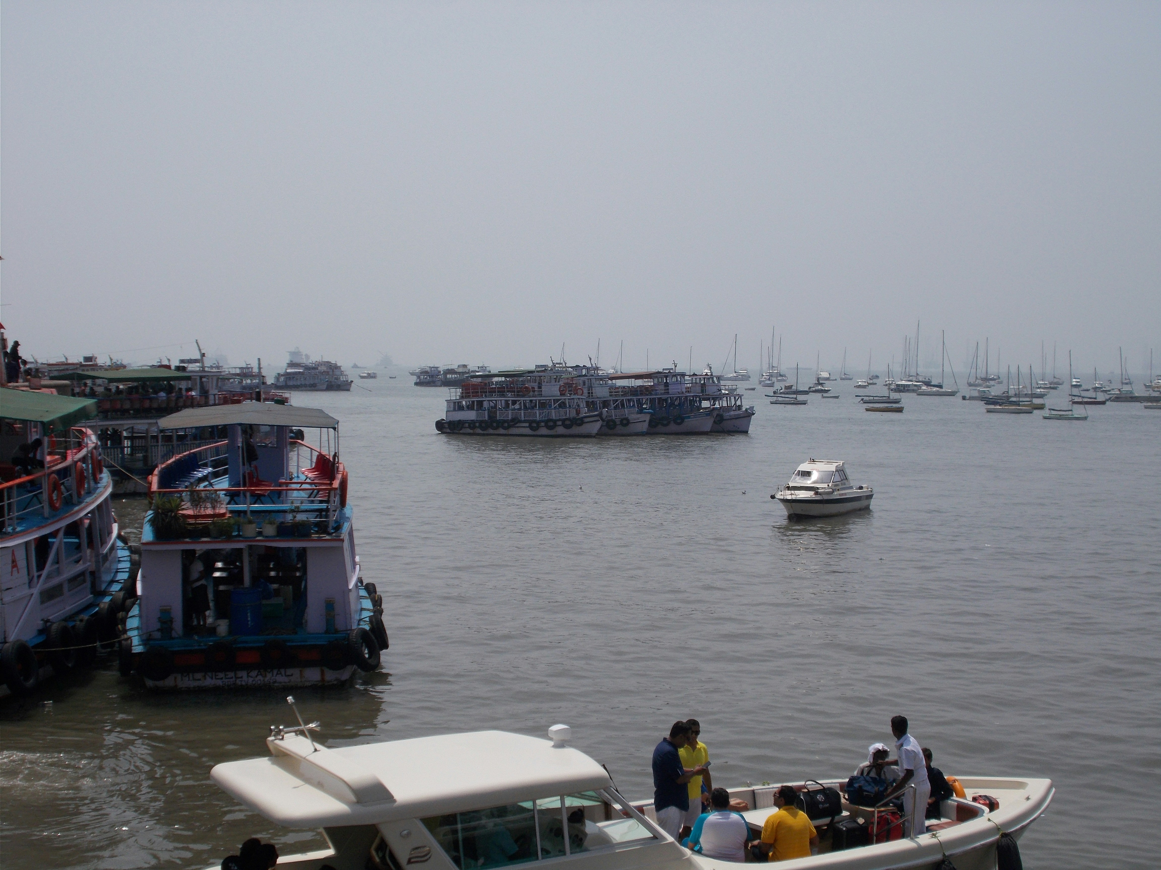 A harbor scene with several boats docked along the quay and a small motorboat in the foreground, while a hazy array of larger vessels fills the background.