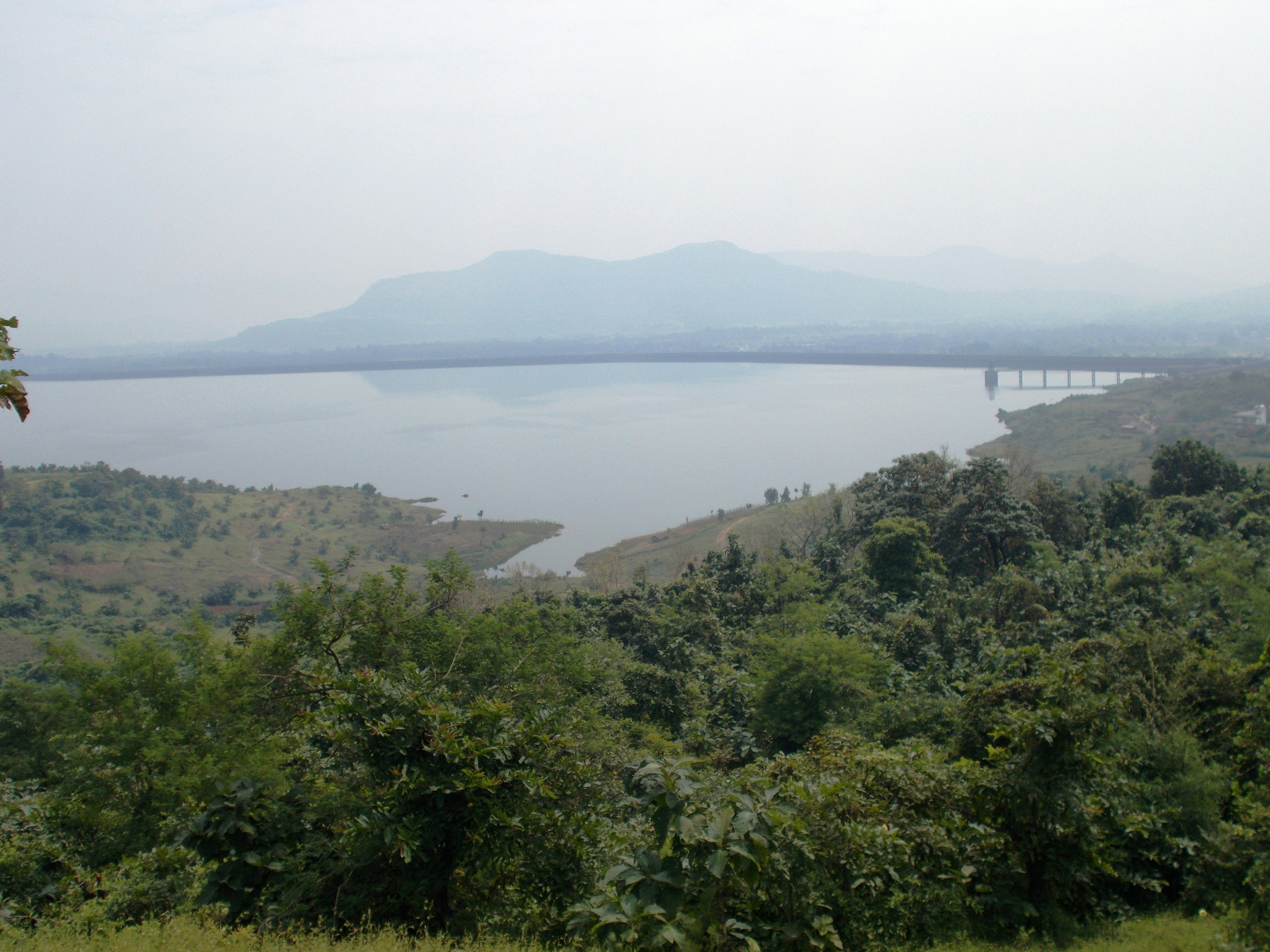 Maintained greenery in the foreground overlooks a calm reservoir with a distant bridge and faint mountain range under a pale sky.