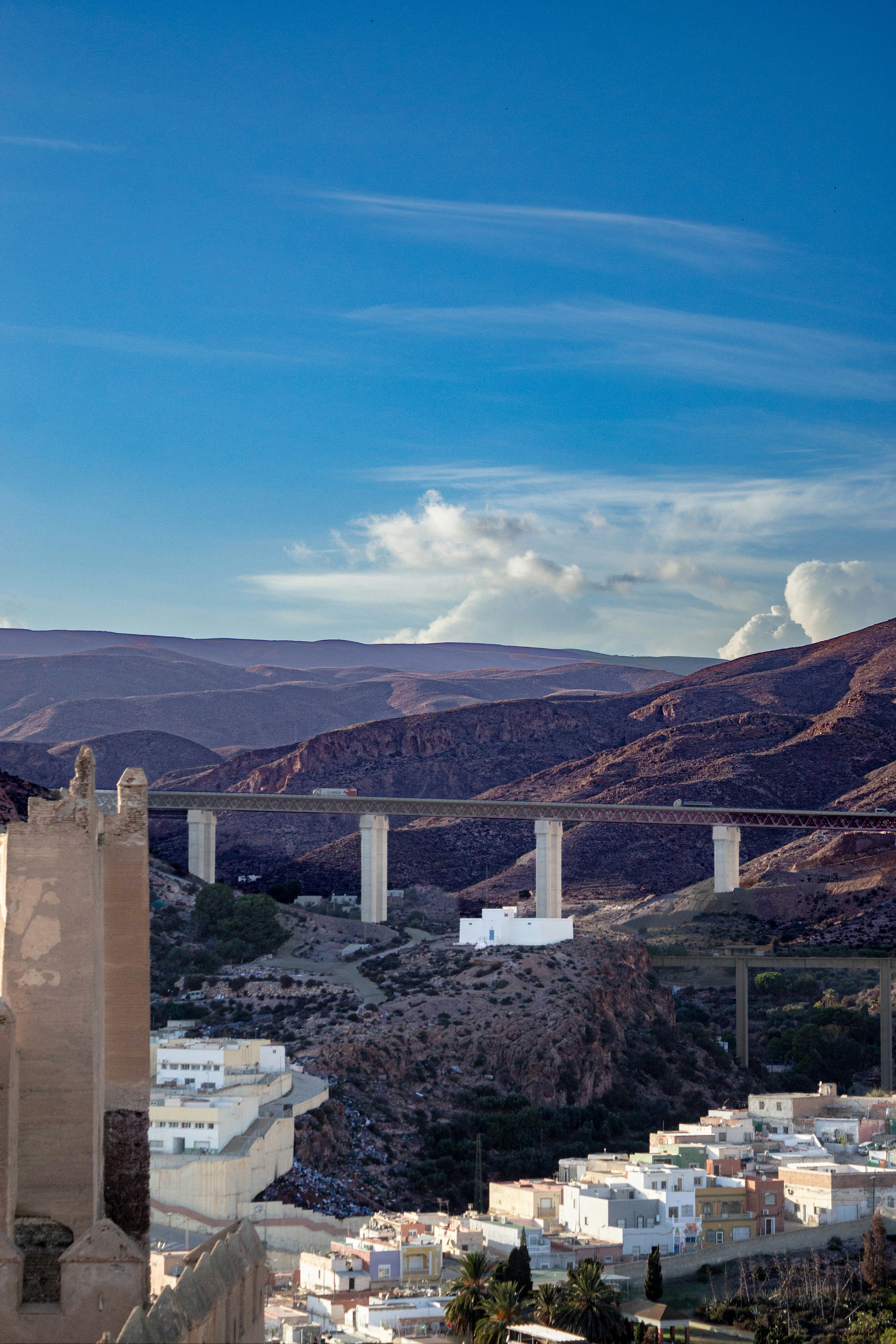 Paisaje de un valle rodeado de montañas áridas. Un gran puente de hormigón cruza el barranco conectando ambos lados del relieve. En la parte inferior aparecen edificios blancos y estructuras urbanas, mientras el cielo presenta nubes alargadas que contrastan con el intenso azul.