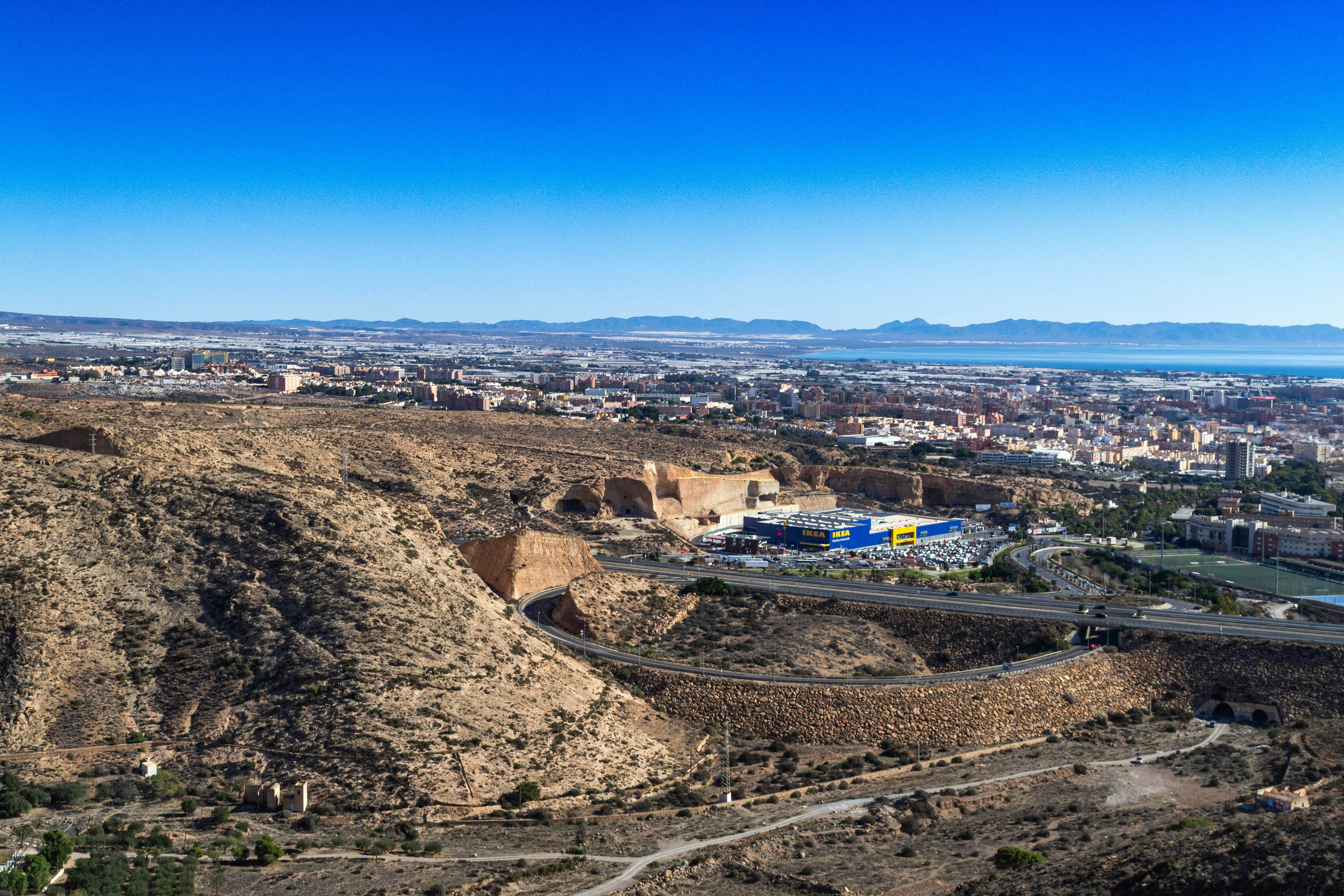 Sunlit hillside leads to a sprawling urban area beyond arid ridges; a winding road threads through the foreground toward the city. The scene captures a contrast between rugged terrain and developed landscape.