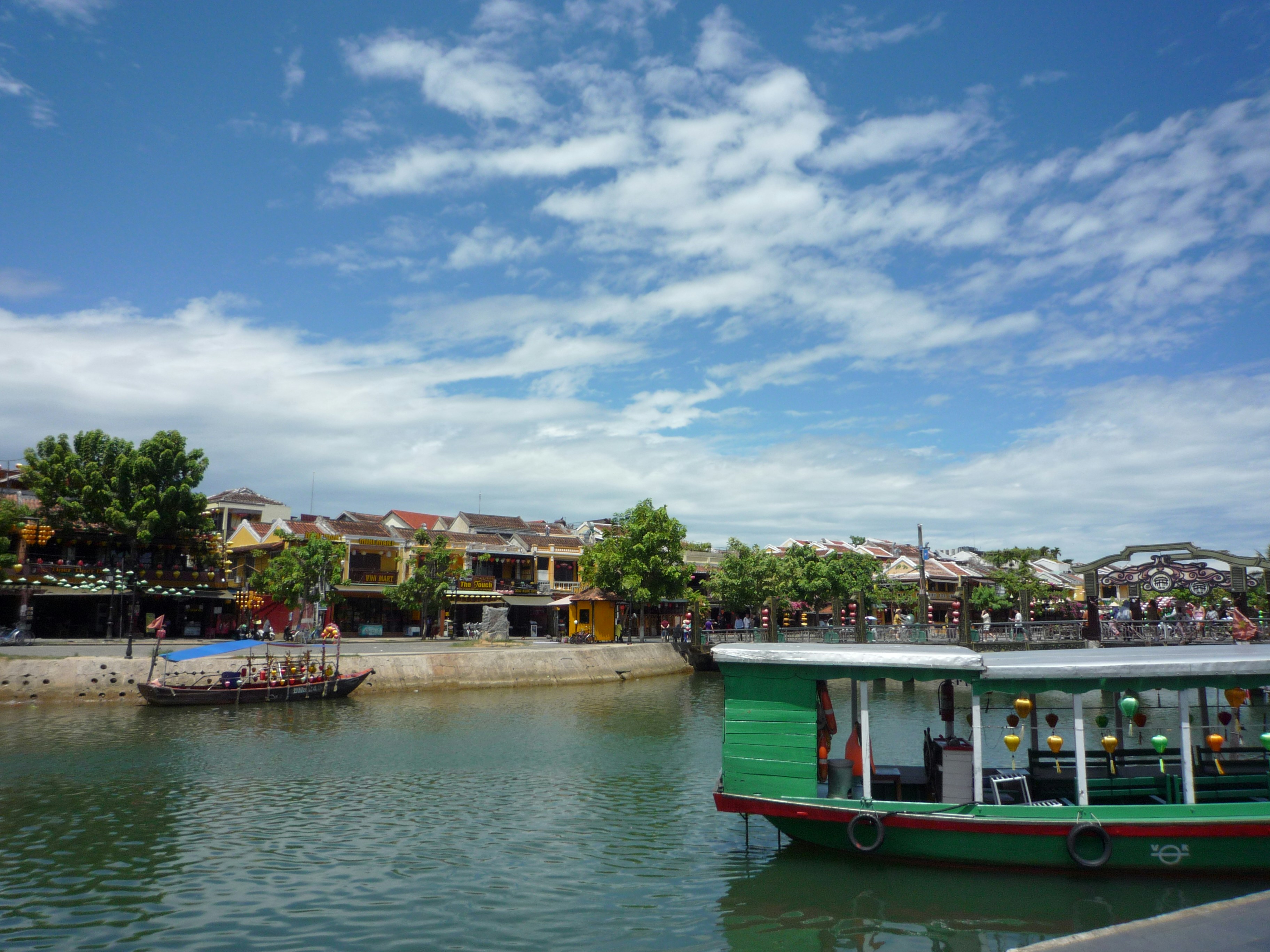 Sunlit riverfront with a green passenger boat in the foreground and a row of colorful buildings along the quay under a blue sky.
