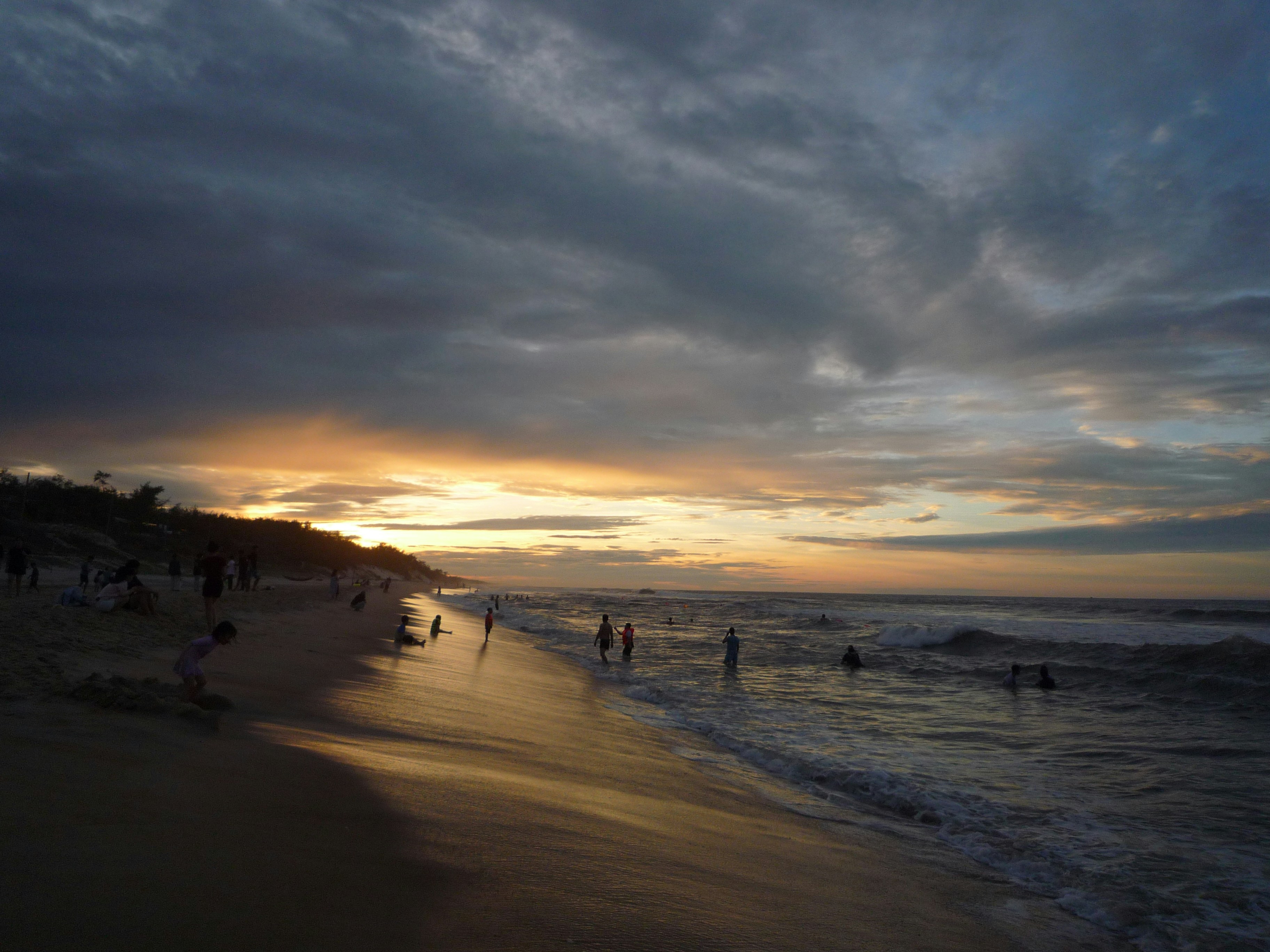 Coastal scene at dusk with people along a sunlit shoreline and calm waves, capturing a tranquil beach moment with a sunset sky.