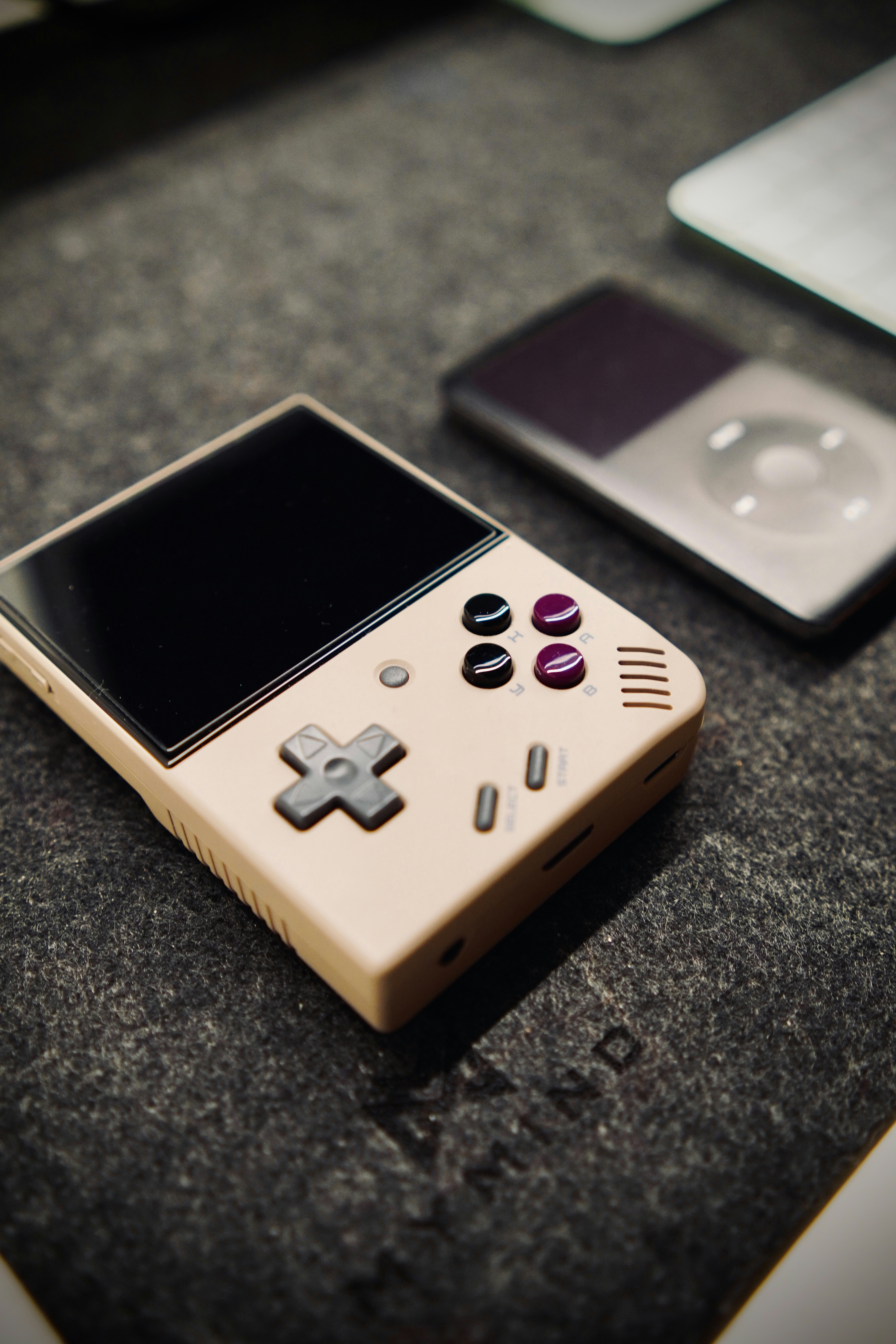 Beige handheld game console resting on a textured tabletop with another device blurred in the background. The photo emphasizes the retro design and tactile buttons.