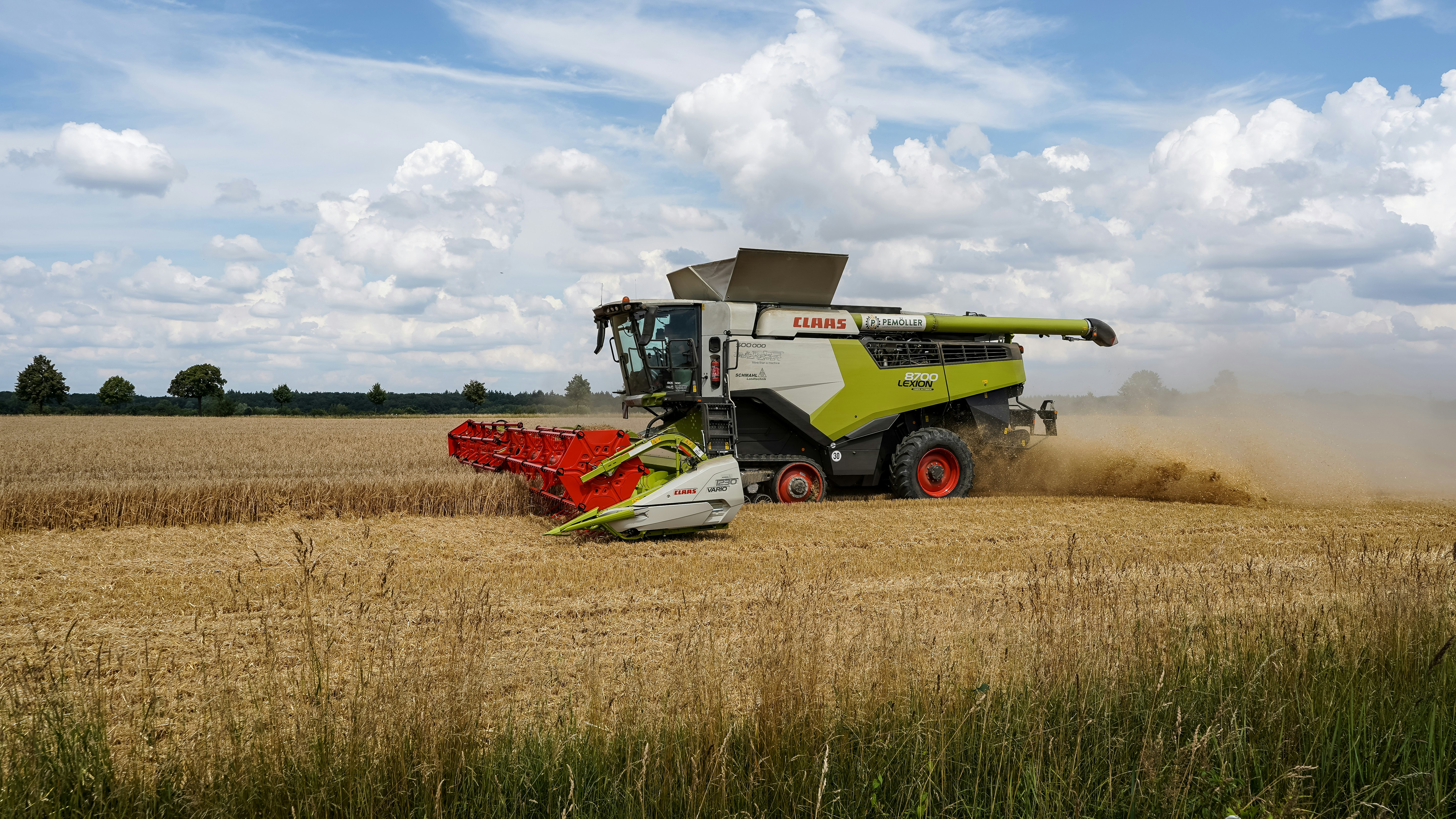 Golden wheat field with a CLAAS combine harvester kicking up dust as it reaps; an expansive rural landscape under a partly cloudy sky.