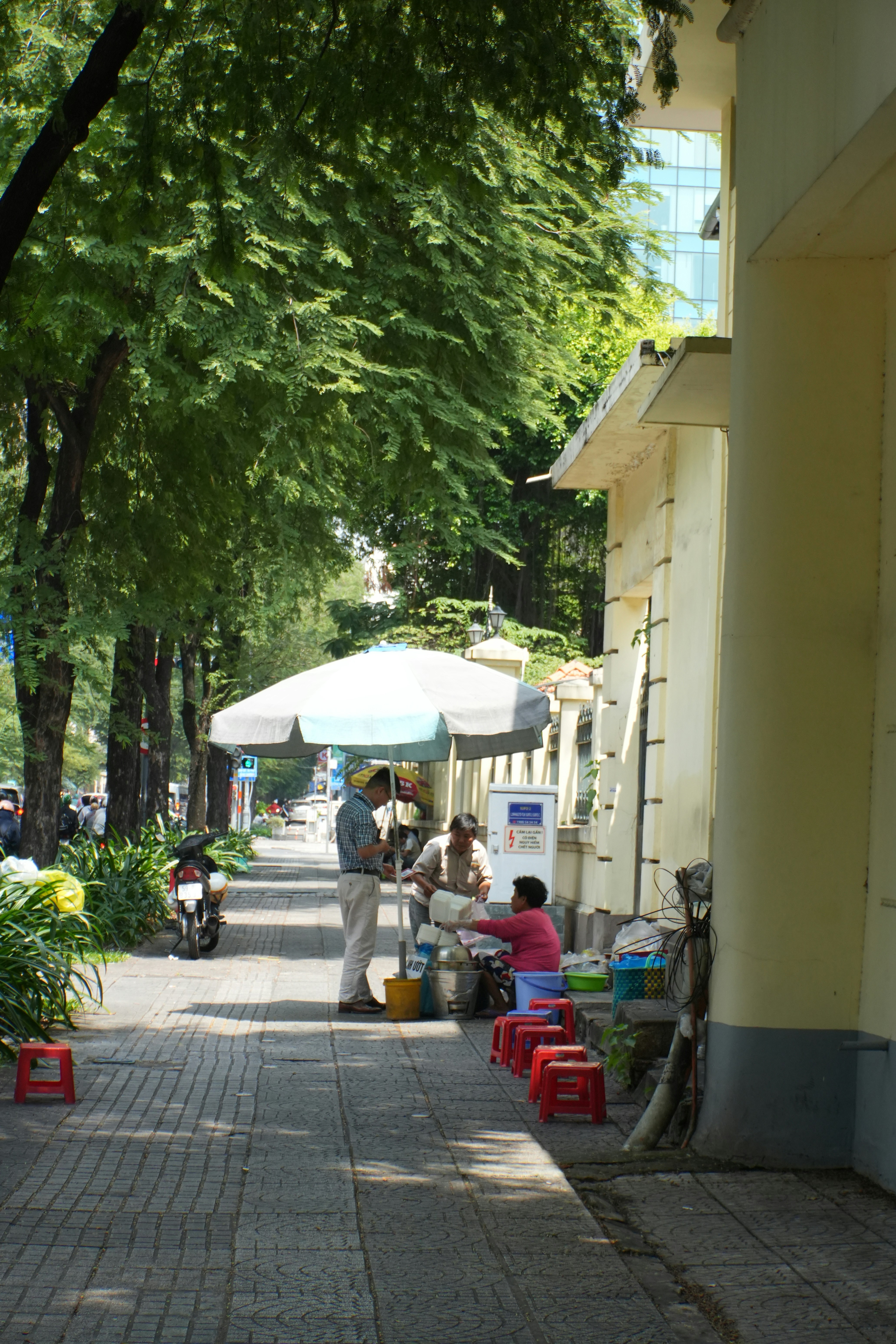 Bustling street scene with vendors under umbrellas along a tree-lined sidewalk; pedestrians converse near a small market setup beside a pale building.