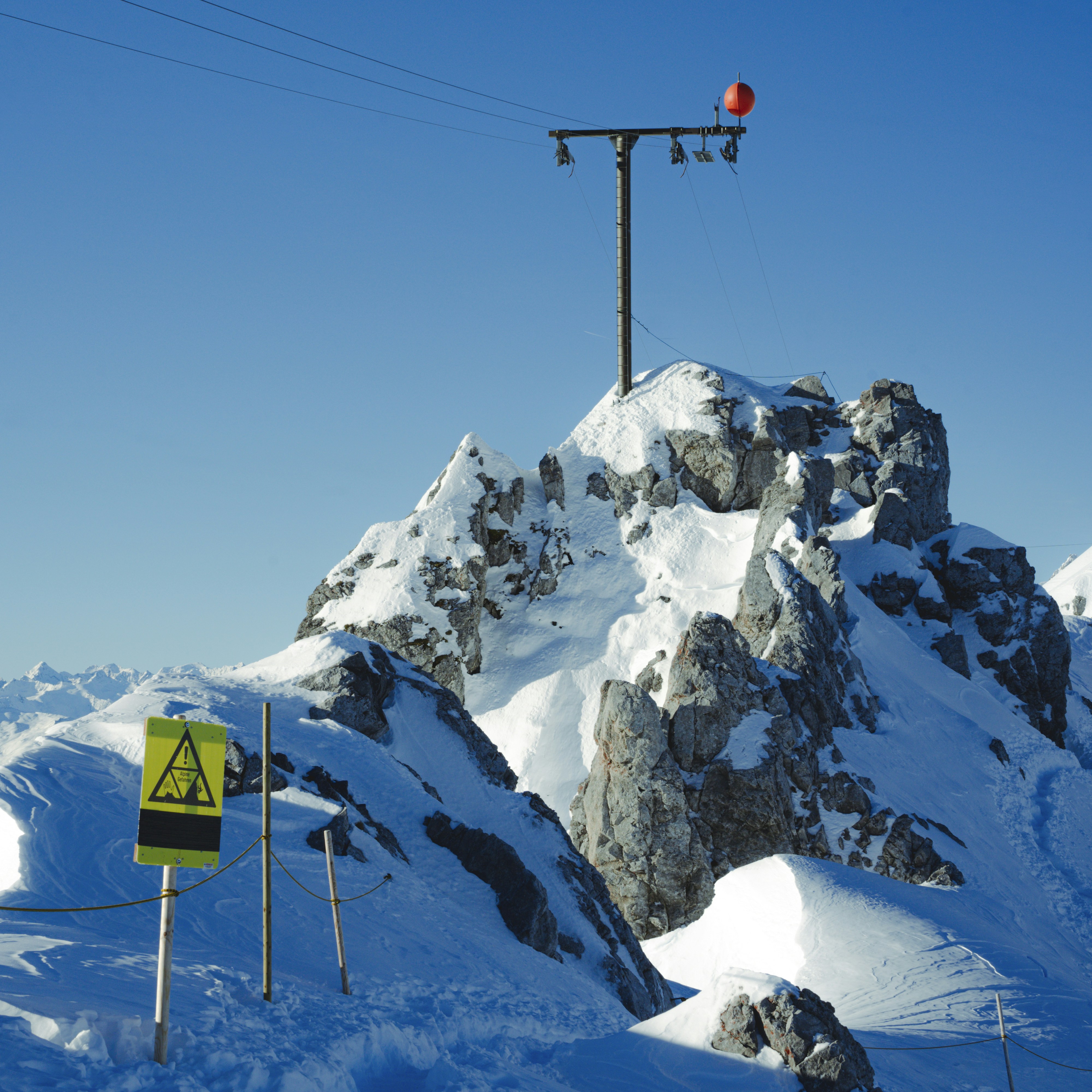 Snowy alpine peak with a cable car support rising above jagged rocks; a yellow warning sign sits at left foreground, all under a clear blue sky.