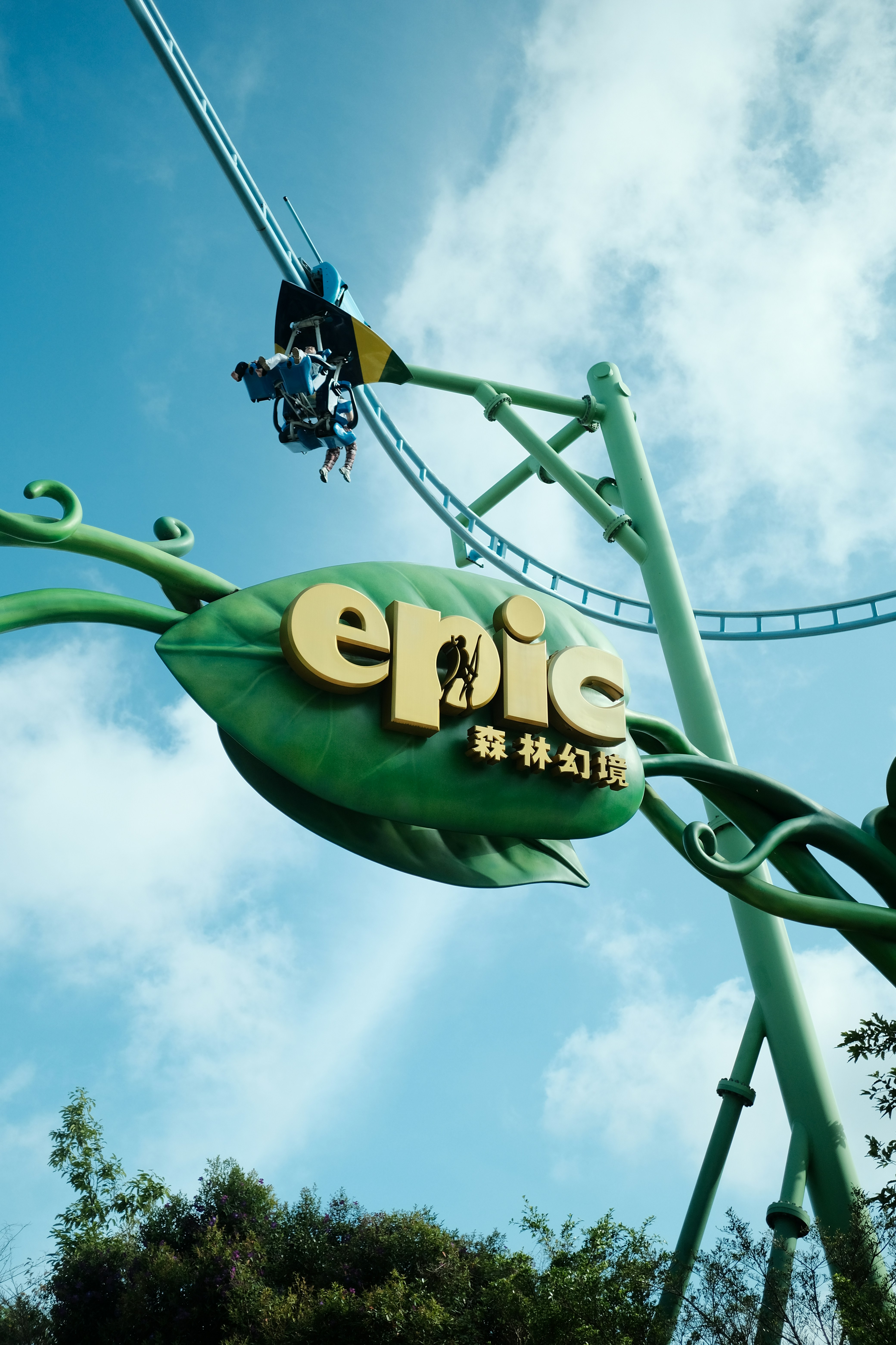 Bright blue sky frames the upper structure of an amusement park ride, with the vibrant green sign and track visible below. Aerial equipment and Chinese characters hint at a themed attraction.