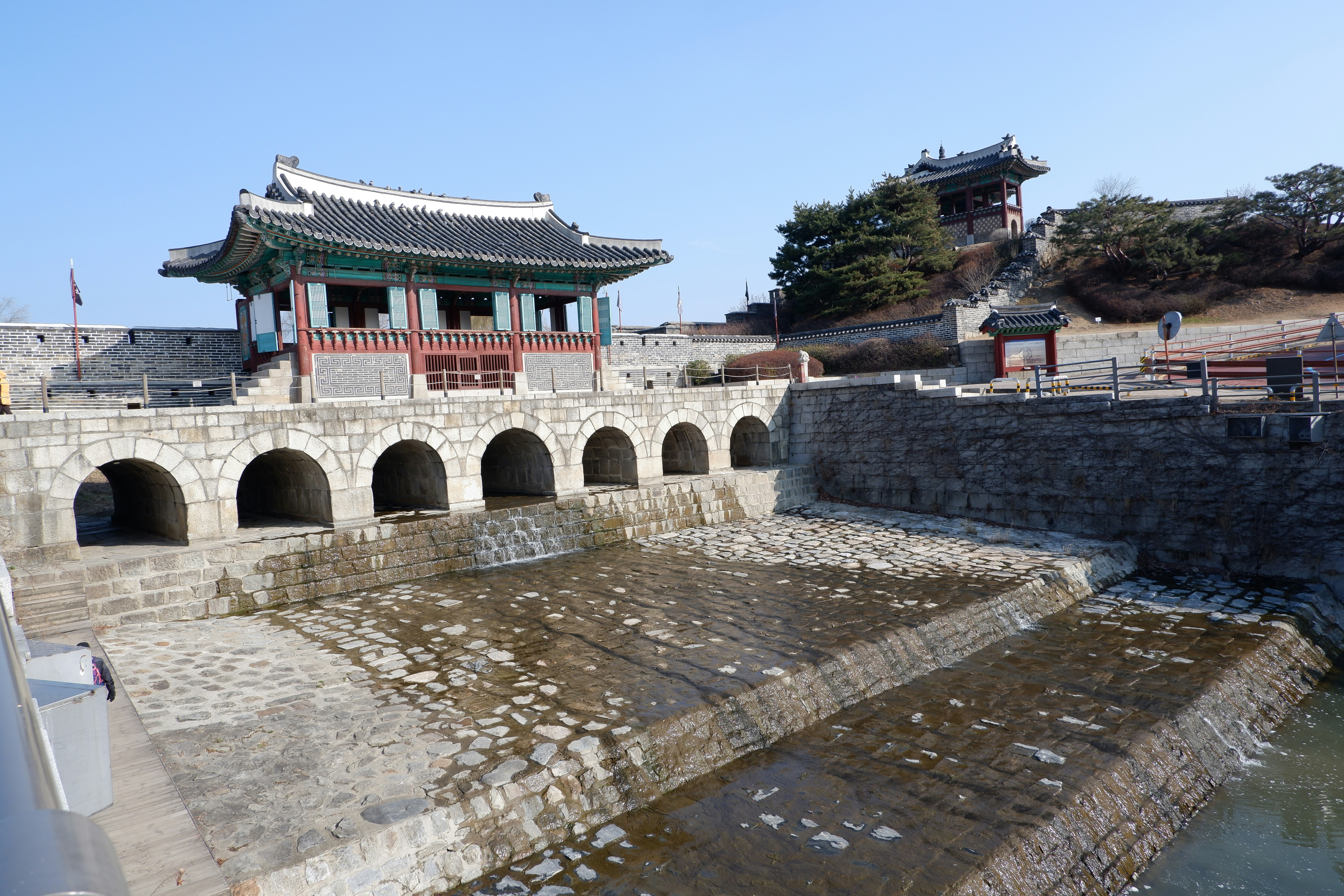 The Beautiful Hwahongmun Floodgate at Suwon Hwaseong Fortress: A scenic and clear daytime view of Hwahongmun Gate, the historic northern floodgate of the UNESCO World Heritage-listed Suwon Hwaseong Fortress in Suwon, South Korea. The photograph beautifully captures the architectural grandeur of the traditional wooden pavilion sitting atop seven structural granite stone archways over the Suwoncheon Stream, with the iconic Banghwasuryujeong Pavilion visible on the hilltop. Perfect for themes on ancient engineering, Korean dynasty heritage, water structures, and cultural travel photography.