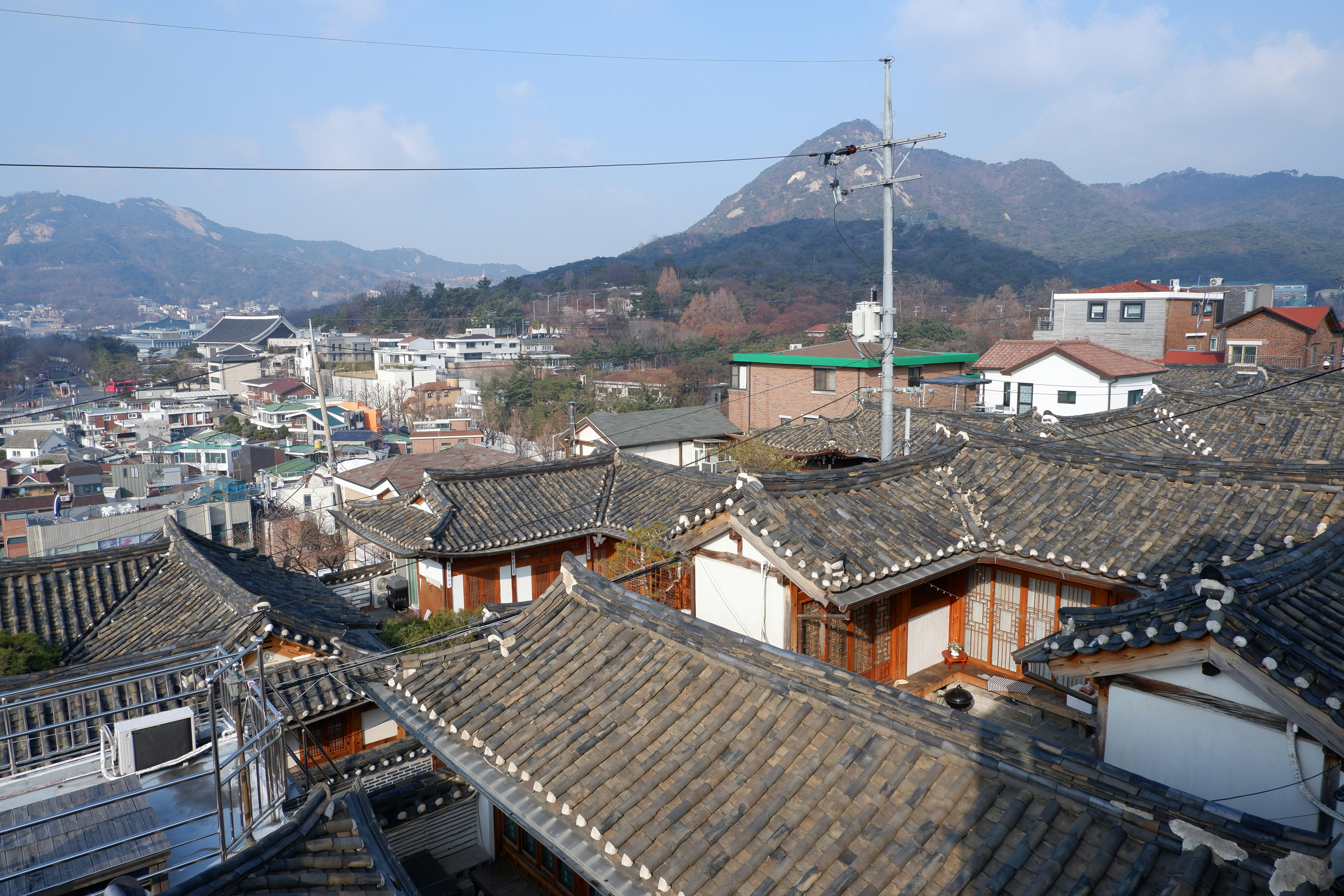 Seoul Cityscape from Bukchon Observatory Rooftop: A stunning elevated view captured from the private rooftop of the famous Bukchon Observatory in Bukchon Hanok Village, Seoul, South Korea. The photo displays a picturesque landscape of traditional Korean tiled Hanok roofs cascading down the hill, contrasting with the residential town below. In the background, the majestic Mount Bugaksan stands tall, overlooking the historical Gyeongbokgung Palace area and Cheongwadae (The Blue House). Perfect for travel memories, cultural heritage, and South Korean tourism collections.