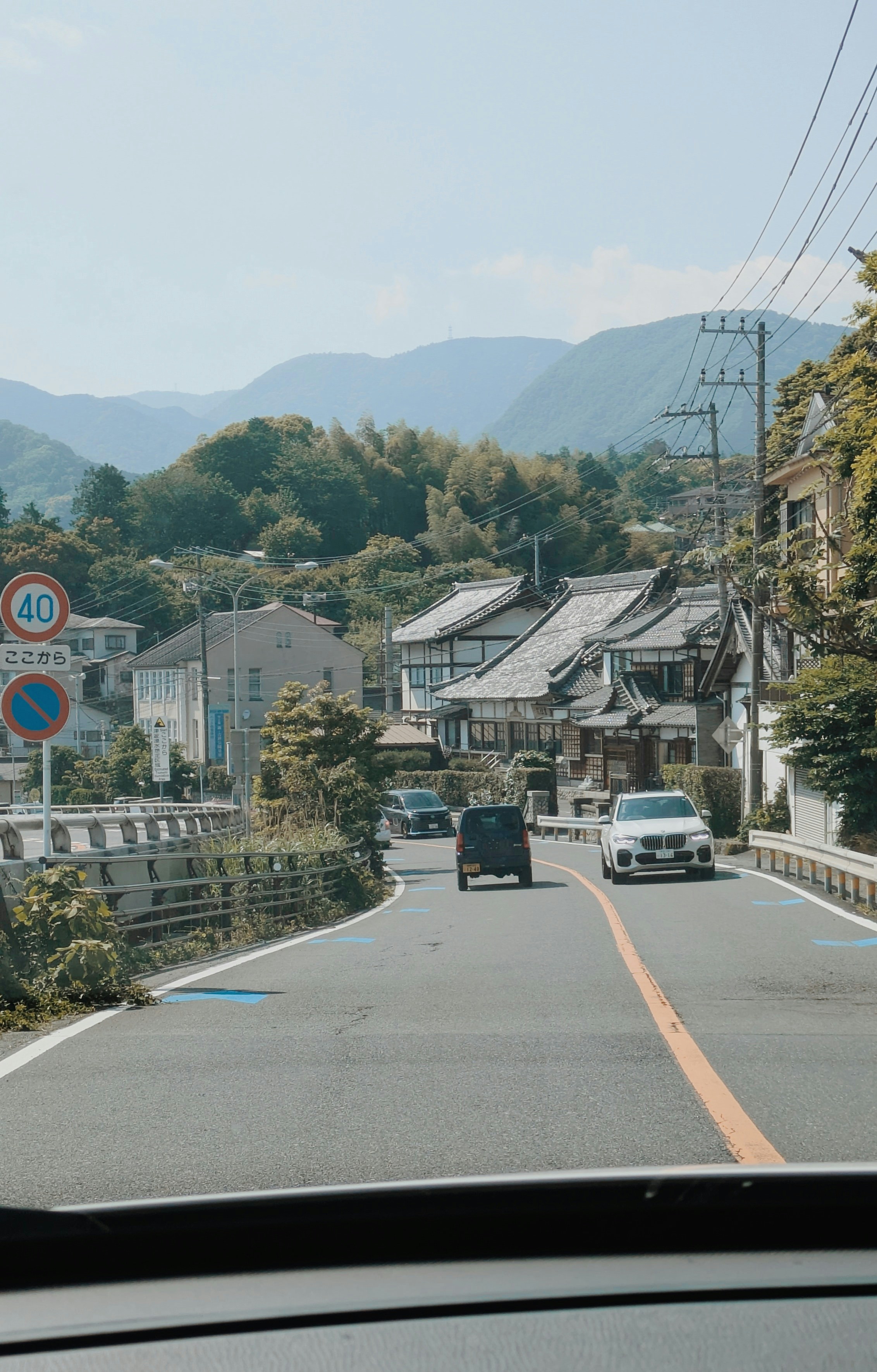 A car-filled street winds through a quiet mountain town, seen from the car's windshield with traditional tiled houses and utility lines, under a clear blue sky. A small sign and distant hills anchor the scene.