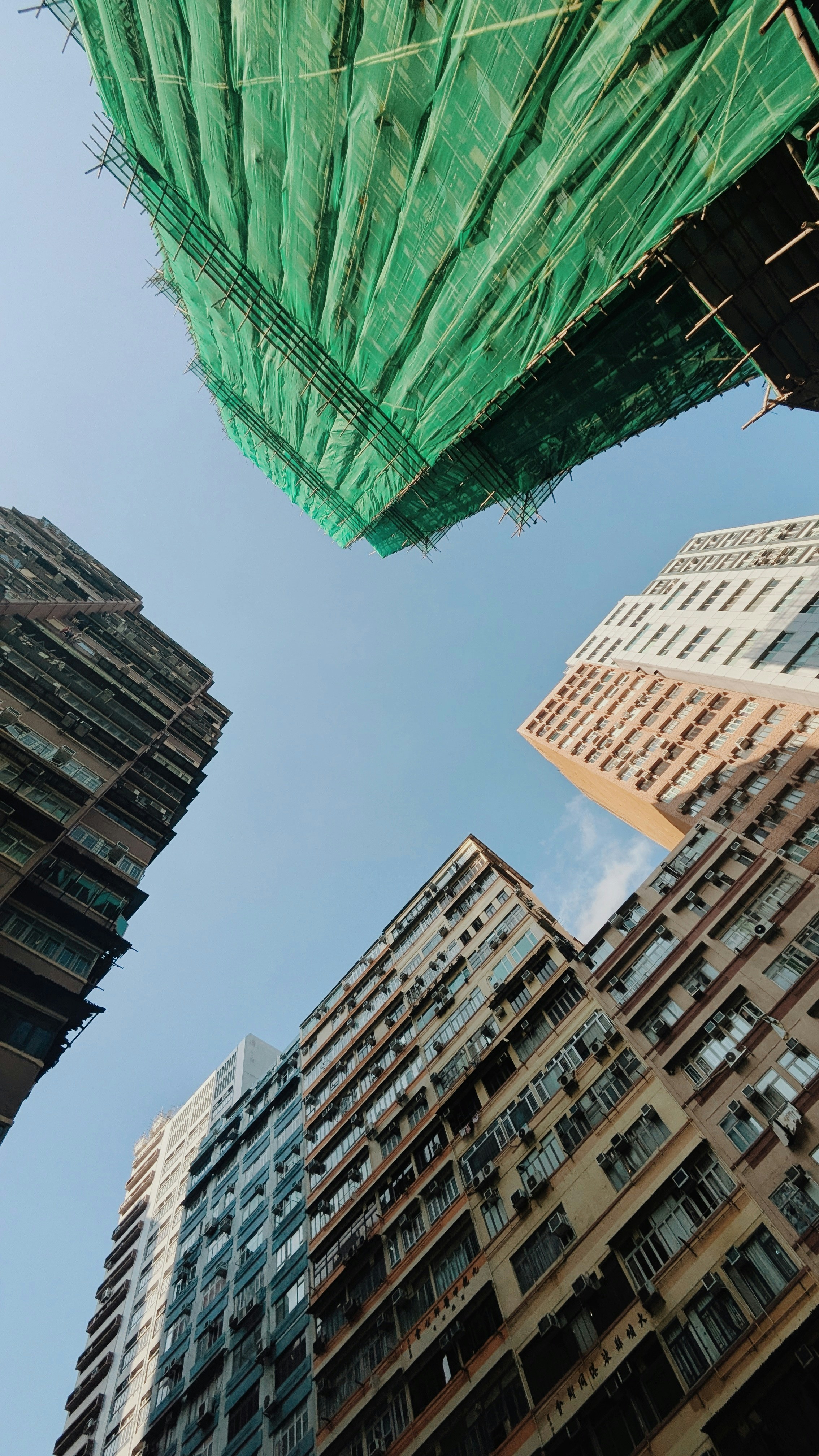 Tall apartment blocks lean inward toward a clear blue sky, framed from a low-angle perspective with a construction-green net at the top. A vertical cityscape snapshot capturing architectural texture and urban scale.
