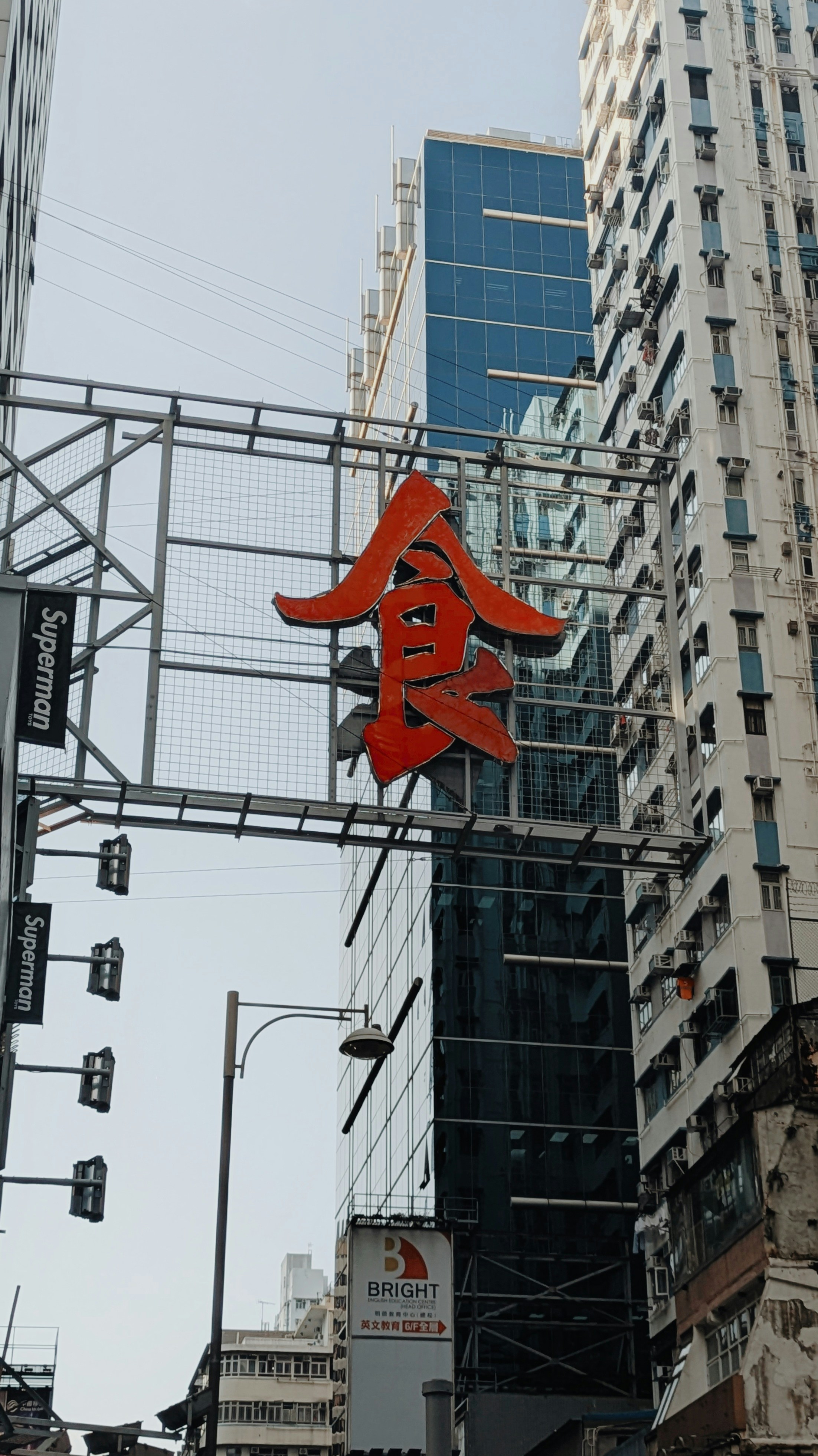 Red Chinese character emblem suspended over a busy city street with modern glass high-rises in the background; a branded sign and scaffolding frame the foreground. Logo-like sculpture stands as the focal point.