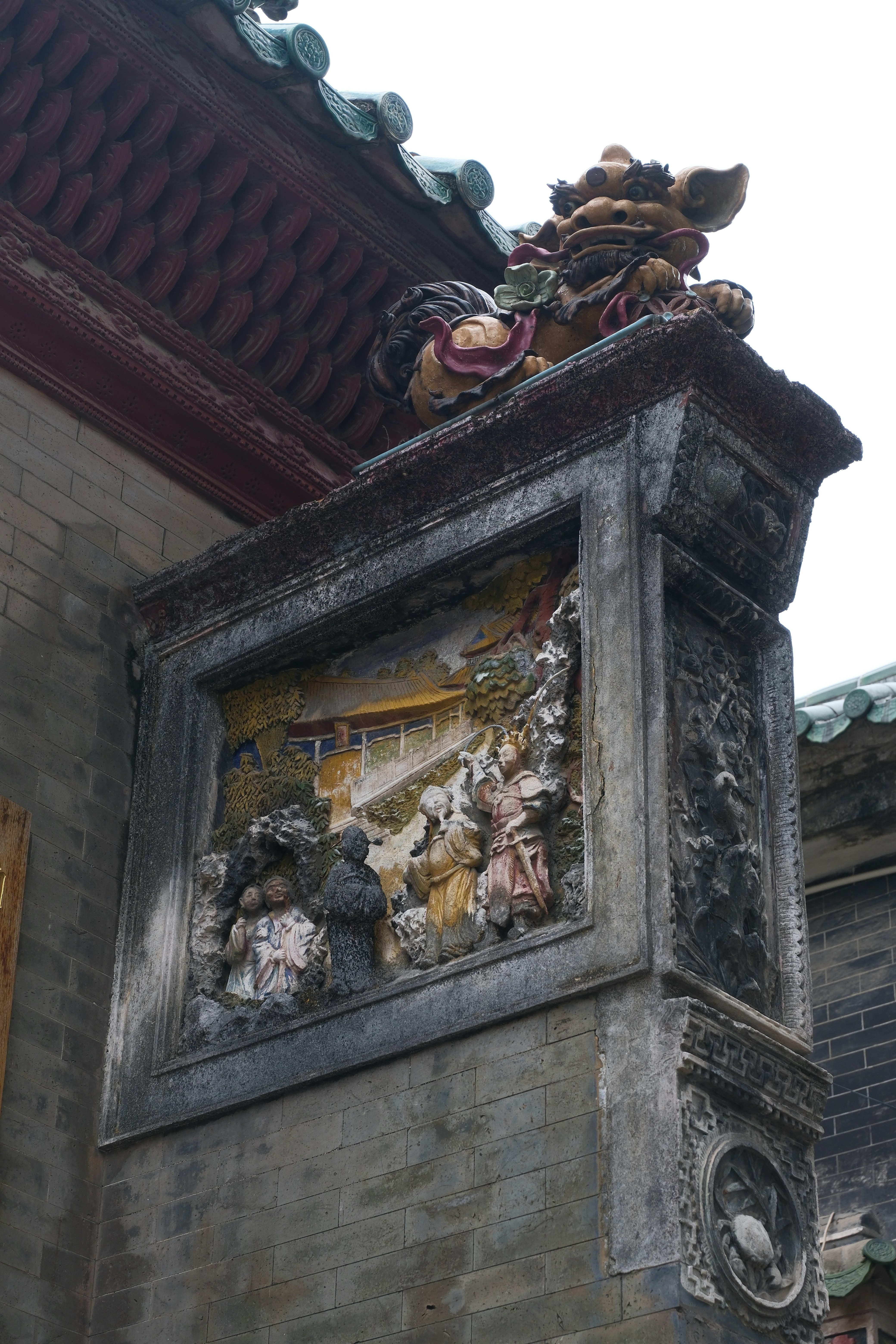 Stone relief with carved figures and a mythical dragon statue atop a temple wall; weathered architectural panel embedded in a traditional Chinese building façade.