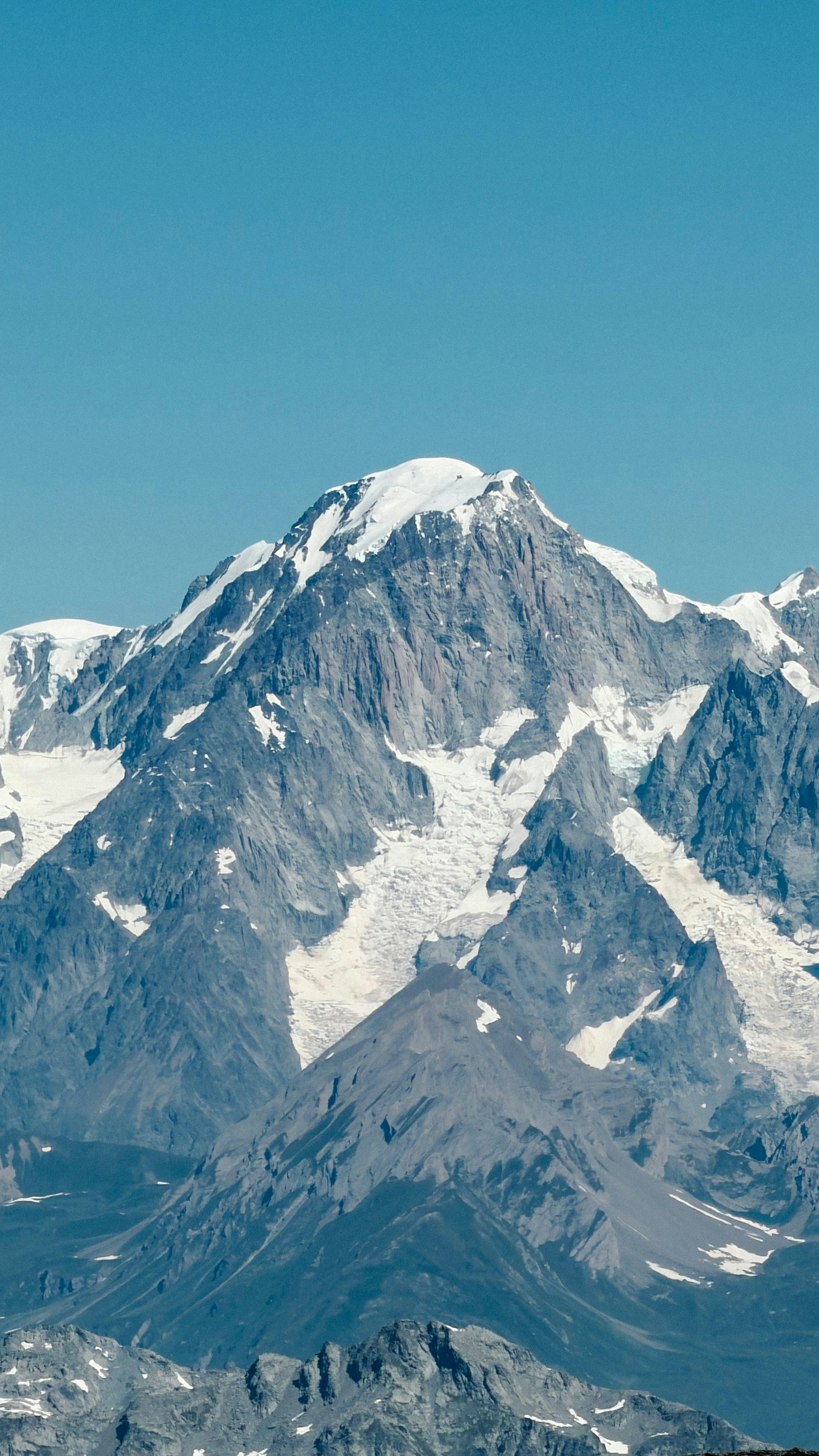 Snow-capped alpine peak rising from rugged slopes against a blue sky, capturing the grandeur of high mountains. A stark, vertical composition emphasizes the ridges and glaciers in cool tones.