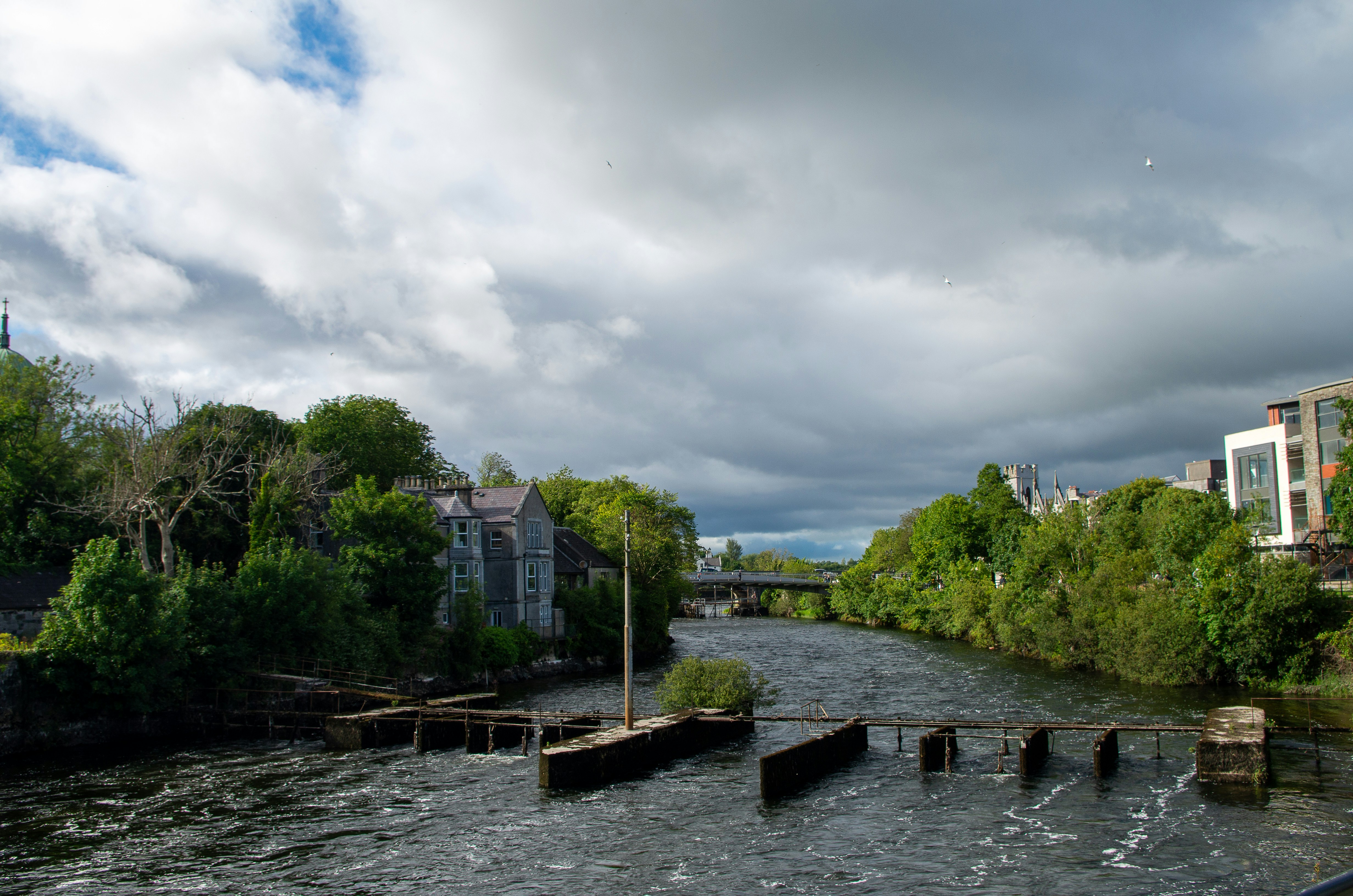 City riverbank under brooding clouds with wooden barriers across the current