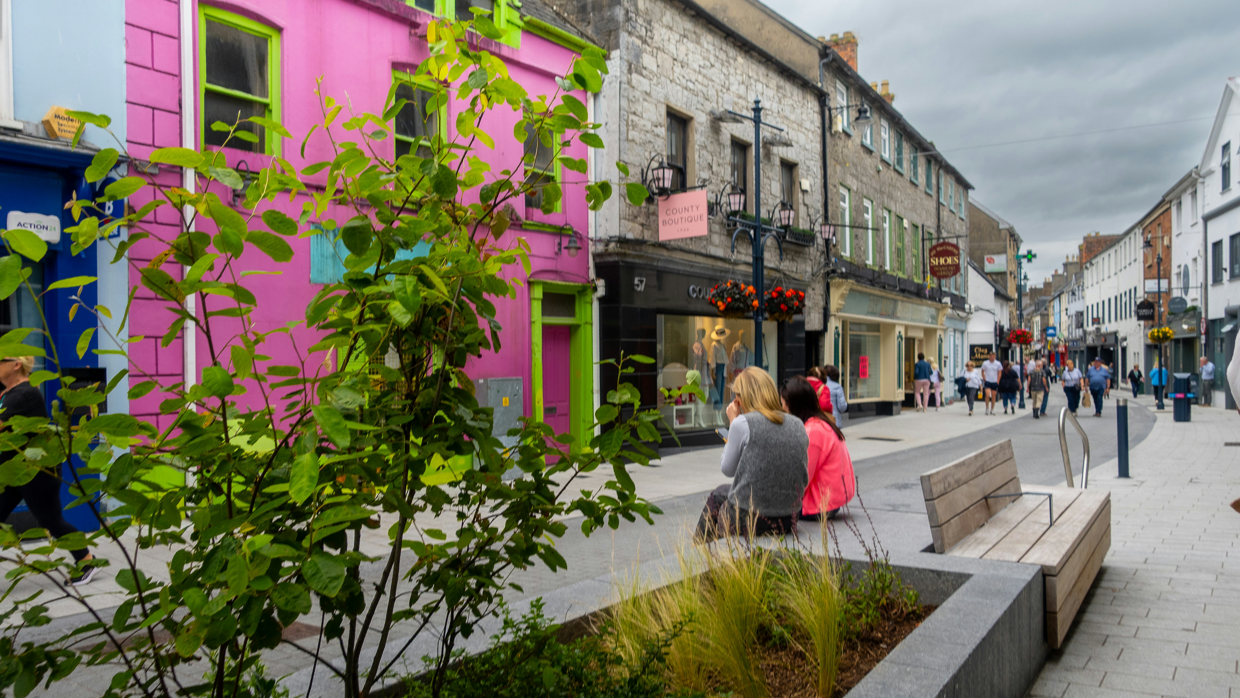 Vibrant storefronts line a pedestrian street while a couple sits on a bench; greenery foreground frames the scene with a busy, cozy urban vibe.