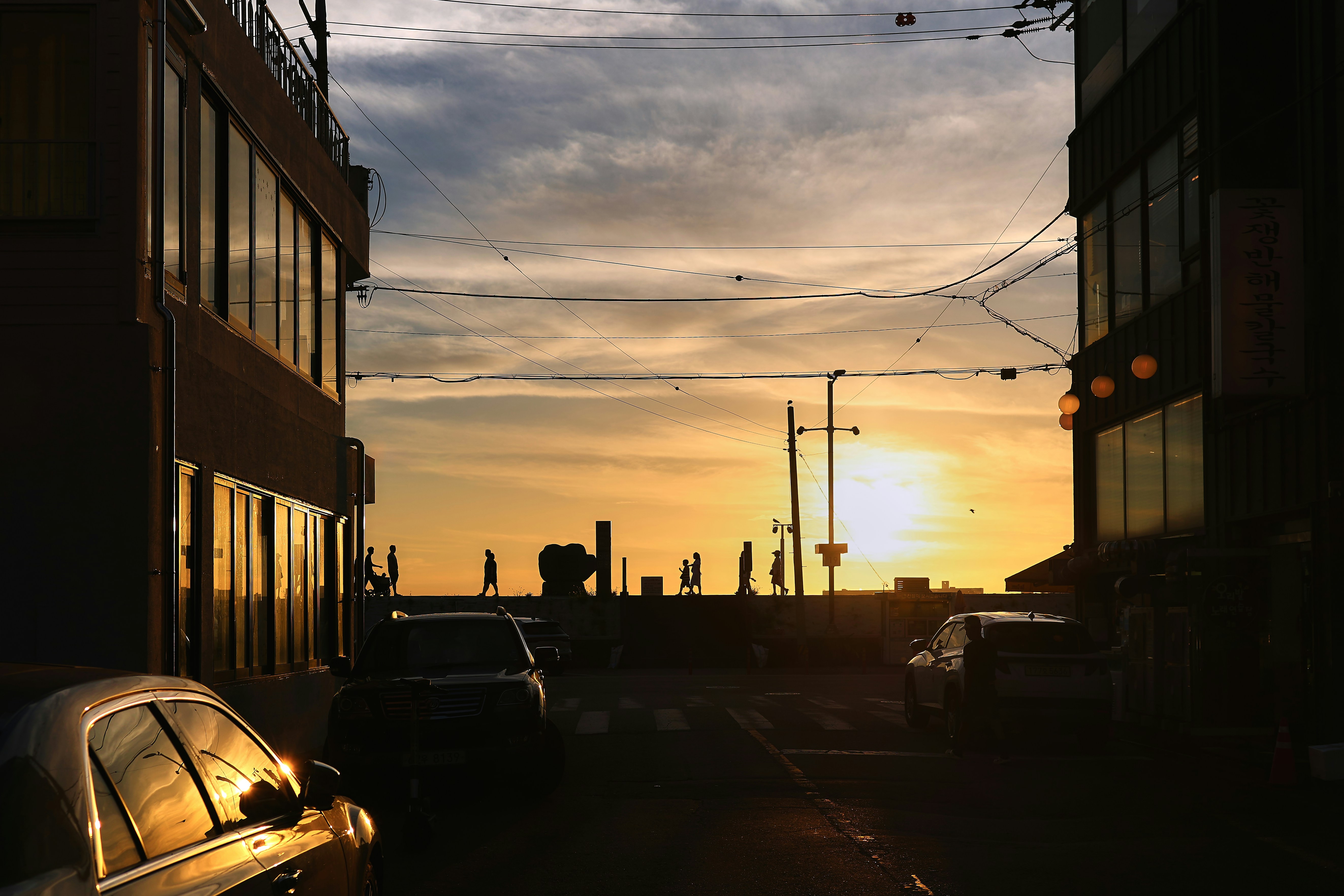 Dusk over a street framed by buildings, with silhouetted figures and parked cars as the sun dips behind the horizon.