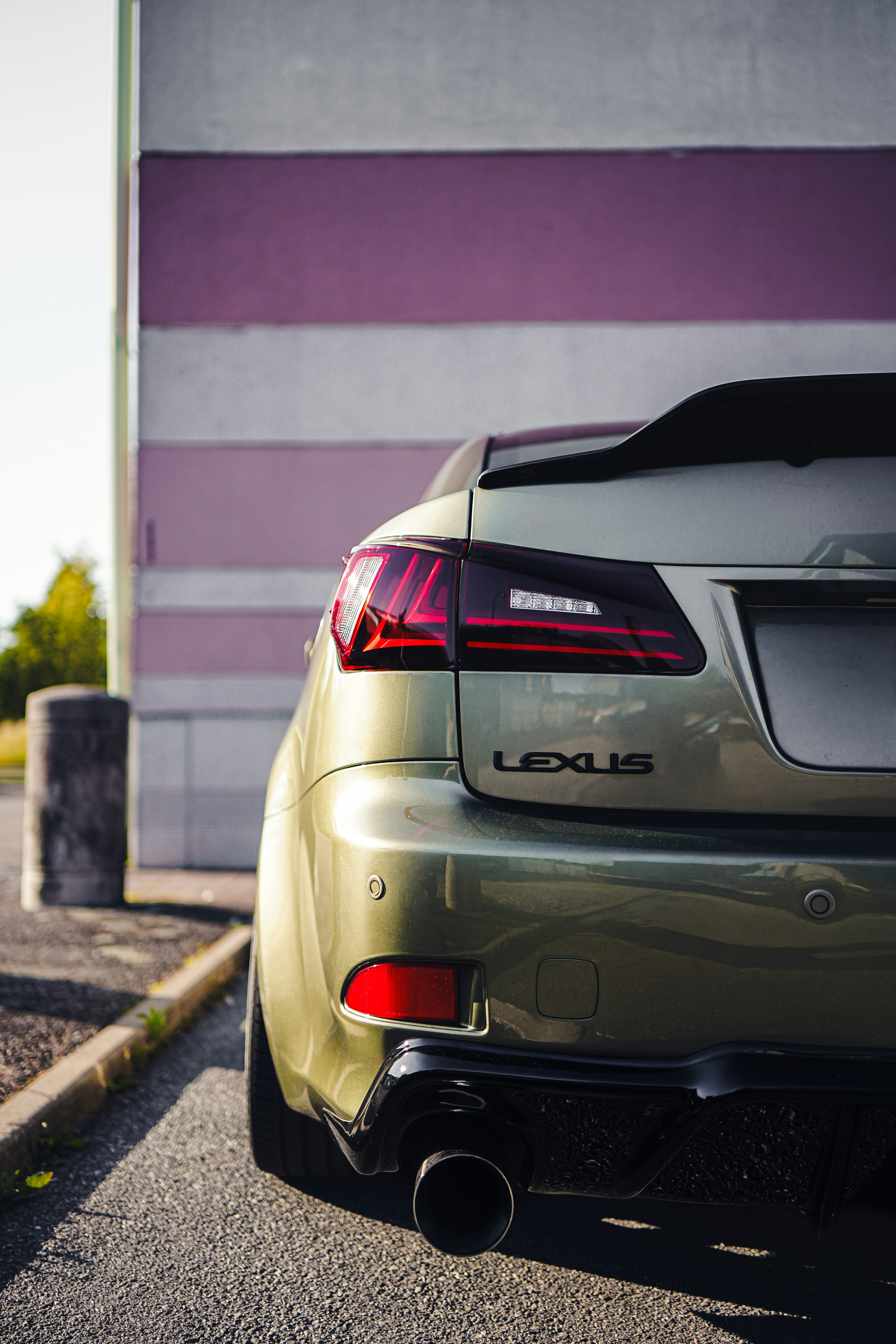 Sleek Lexus rear with distinctive taillight, badge, and exhaust in a sunlit urban setting; a close crop emphasizing the car’s tail and lower bumper. (Photograph is a car shot.)