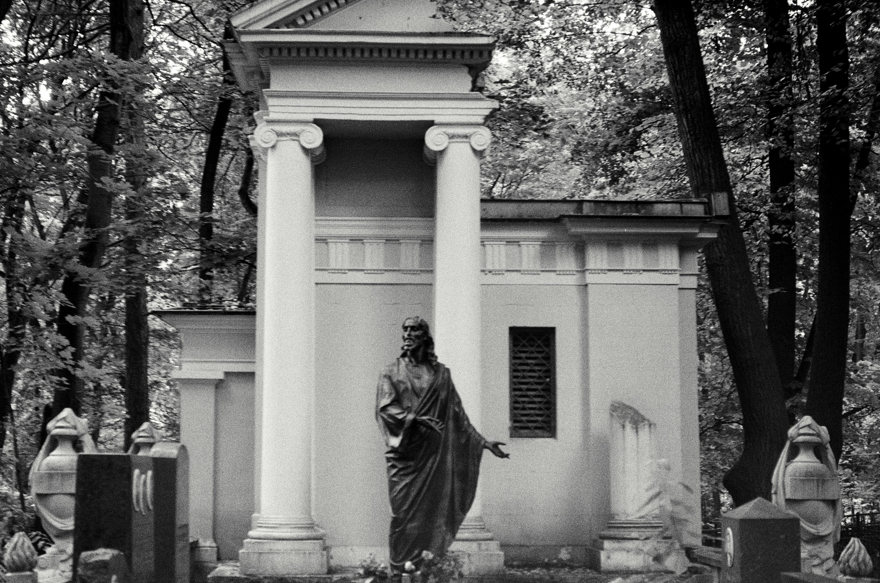 A monochrome scene of a cloaked statue standing before a classical communal tomb, framed by trees and gravestones. The image emphasizes solemn architecture and quiet cemetery atmosphere.