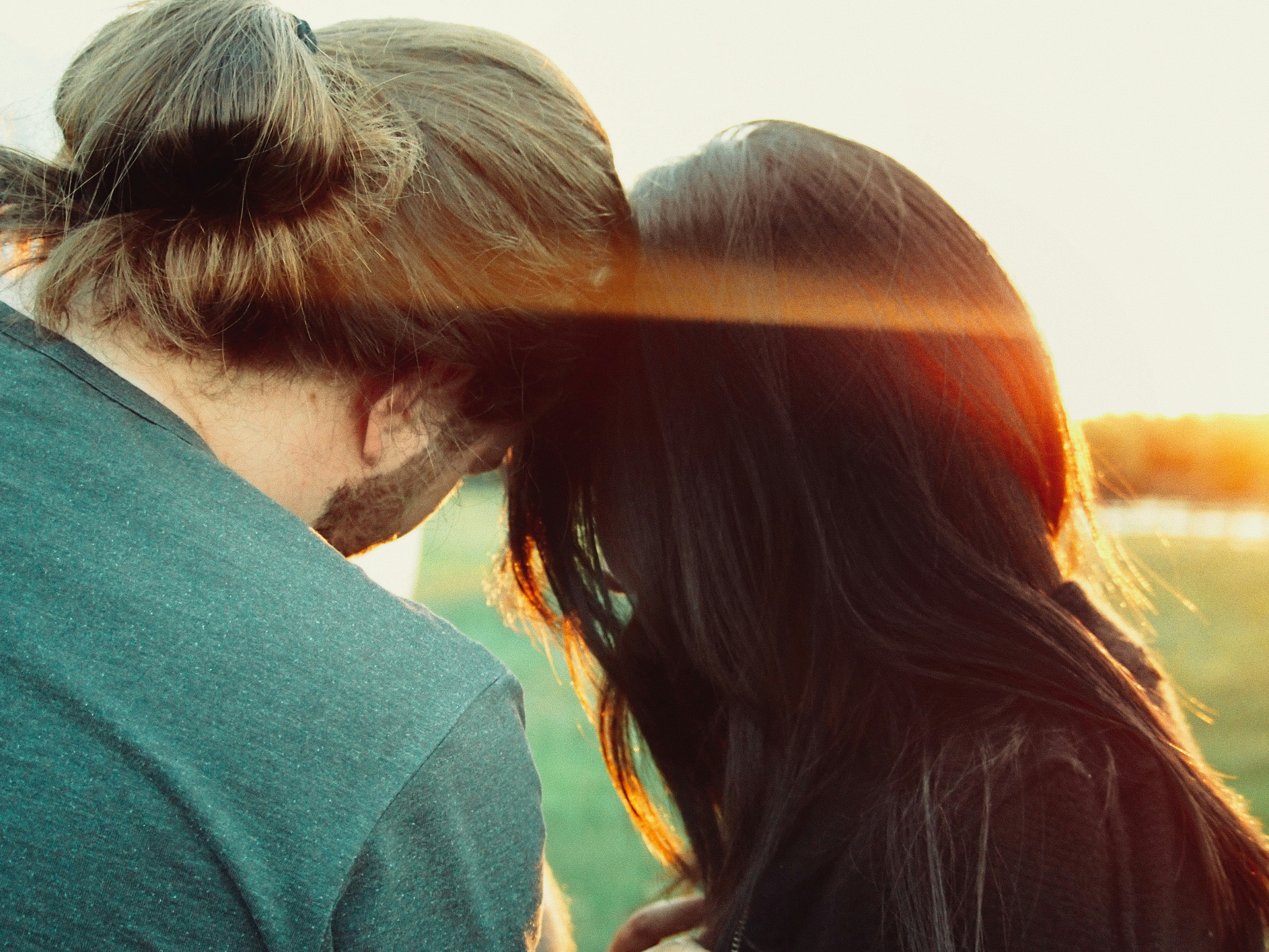 Two people lean in close at sunset, their heads touching as golden light washes over their silhouettes in a sunlit field.
