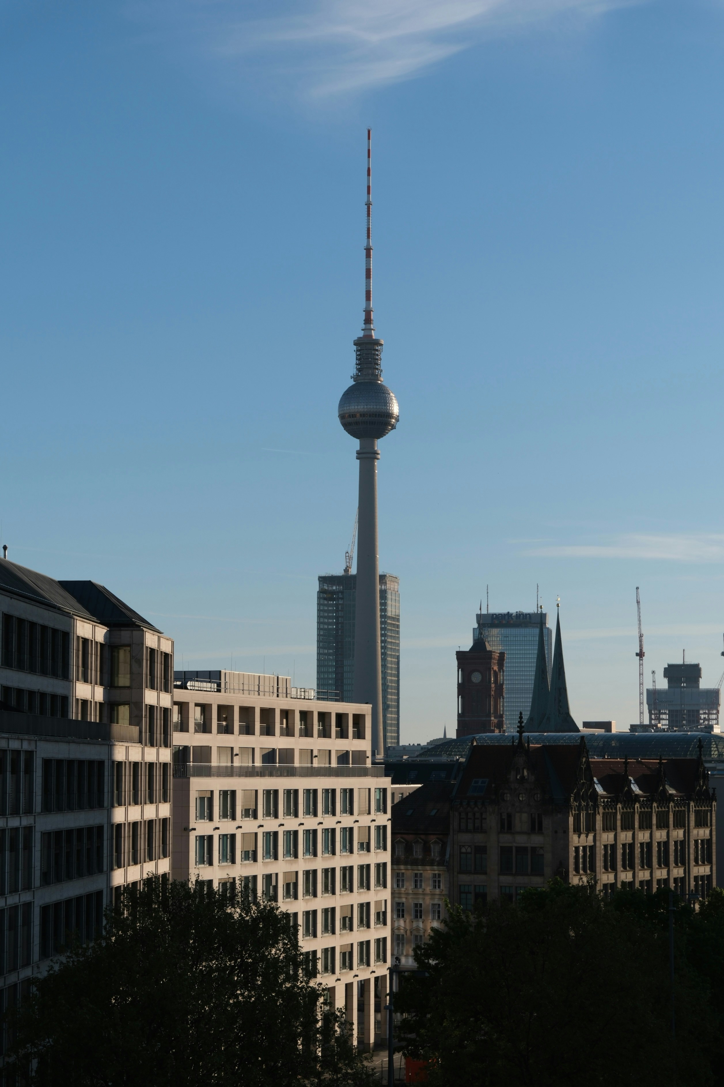 City skyline with the Fernsehturm tower rising above a mix of modern and historic buildings; late-afternoon light casts long shadows across the urban landscape.