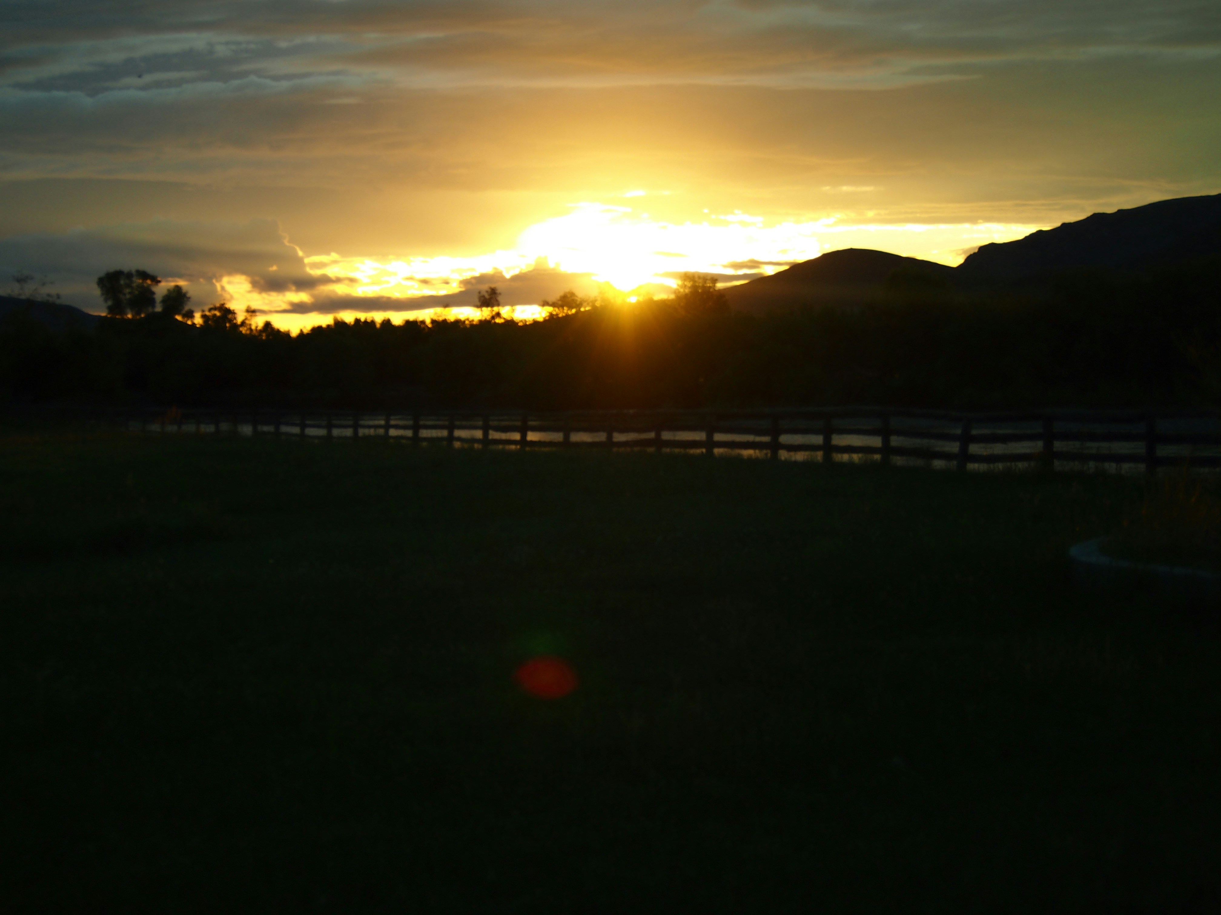 Horizon scene with a setting sun casting warm light over a fence and darkened foreground; a tranquil rural landscape with distant hills.