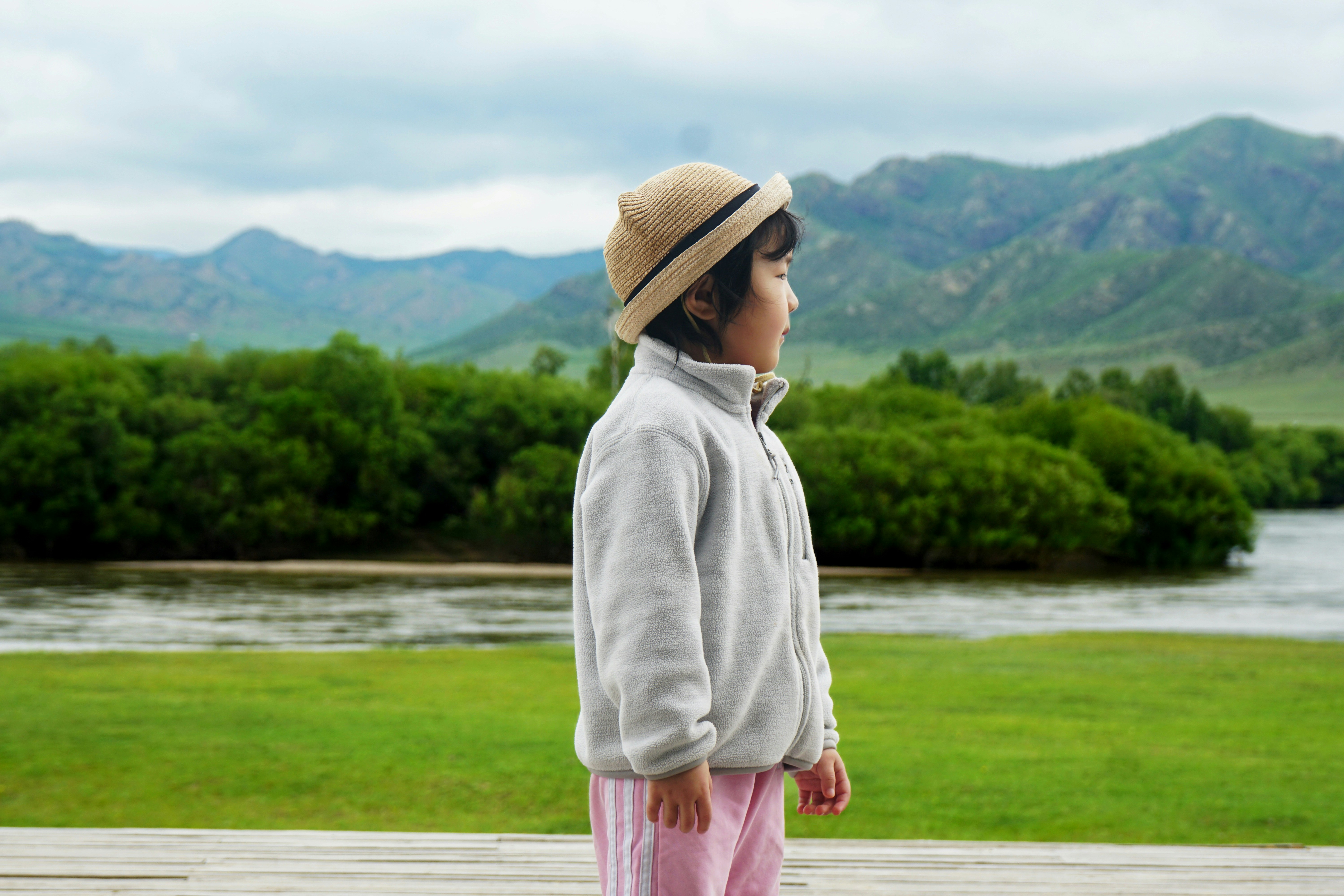 A young child in a light fleece jacket and straw hat stands on a wooden deck by a calm river, framed by distant hills and green shrubbery.