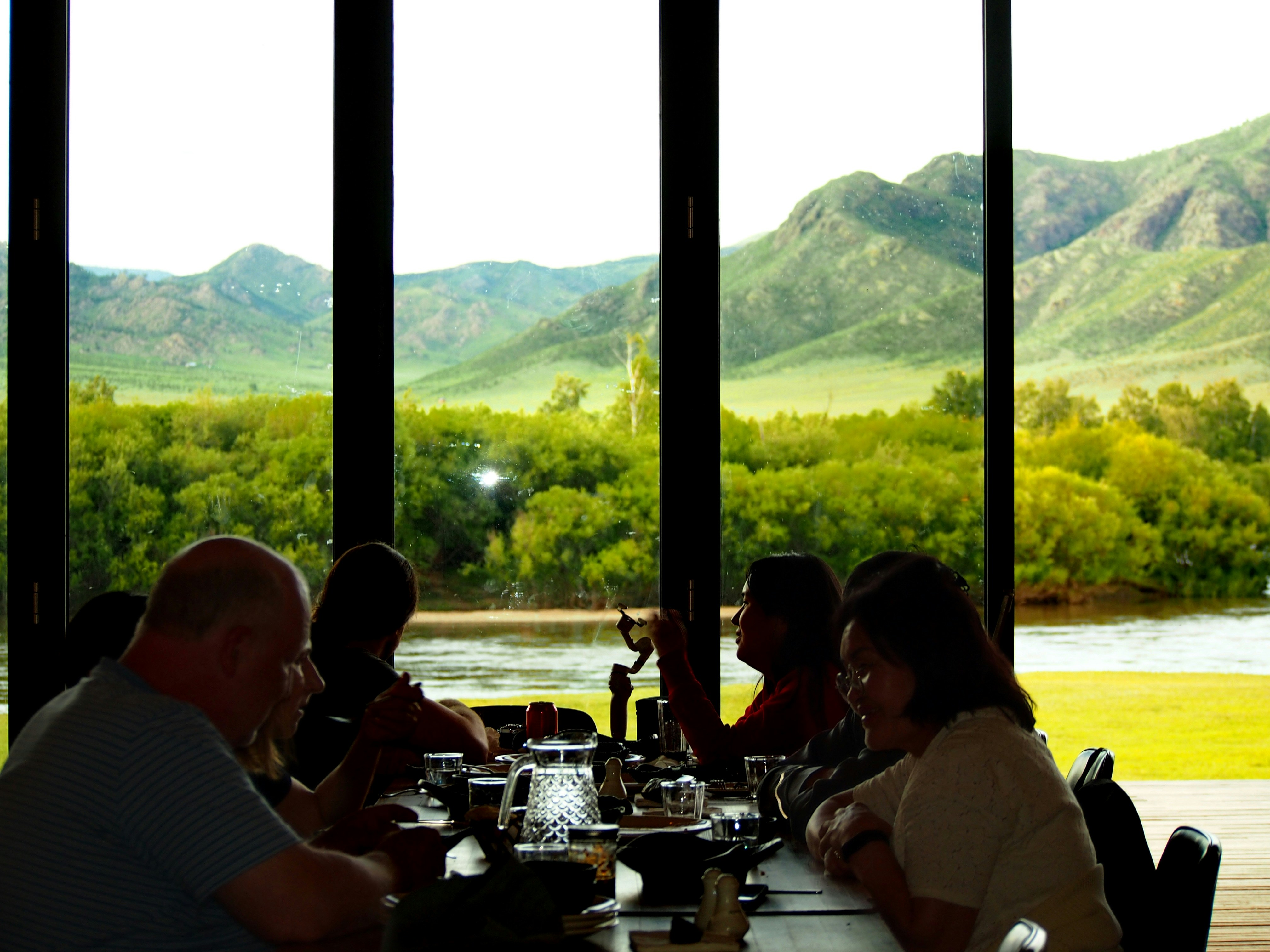 A dimly lit restaurant scene with patrons seated along a long table, dark silhouettes against a bright window showing a river and distant hills.