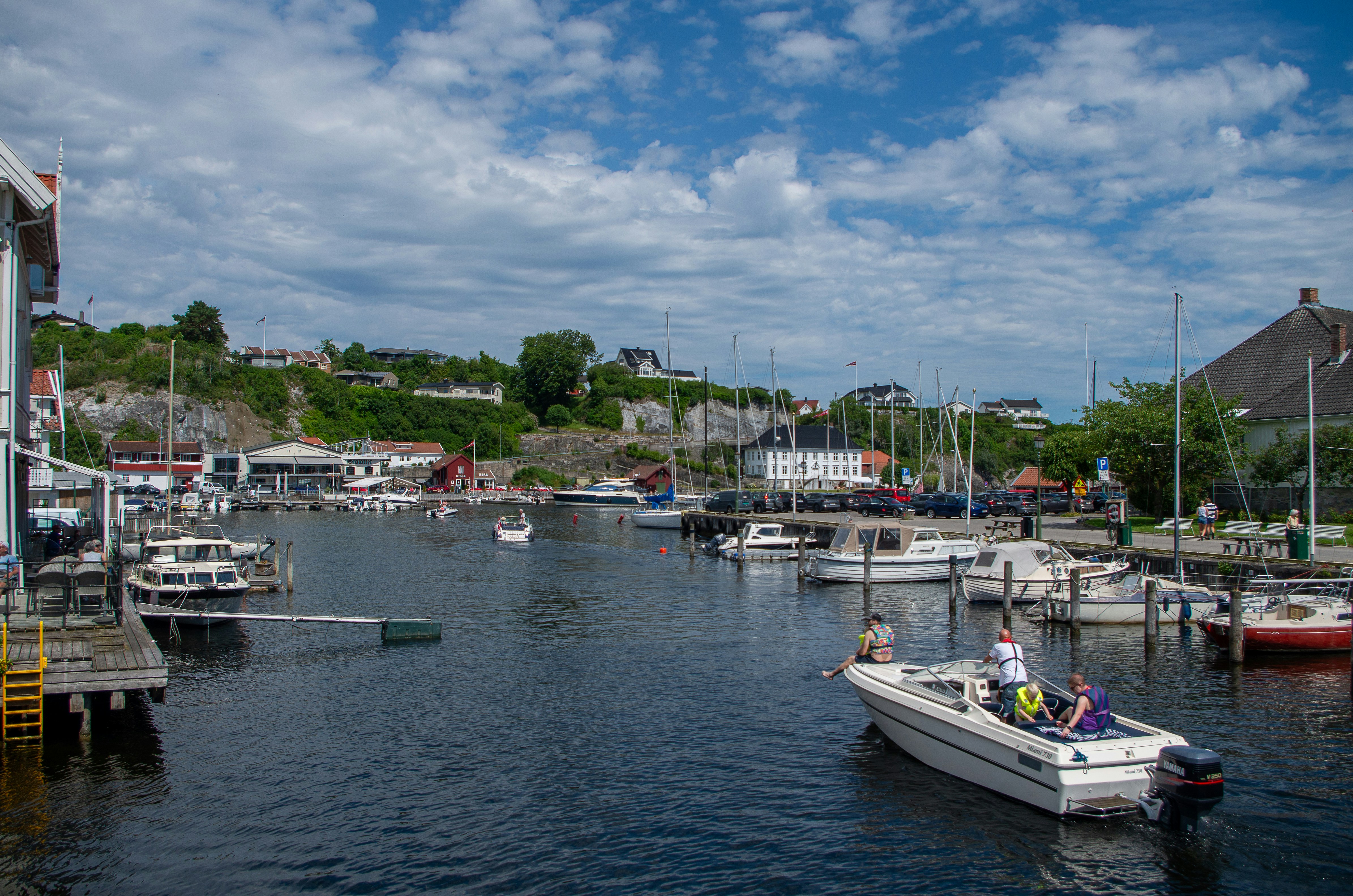 Busy harbor scene with boats docked along a calm waterway and a cluster of houses lining the shore.