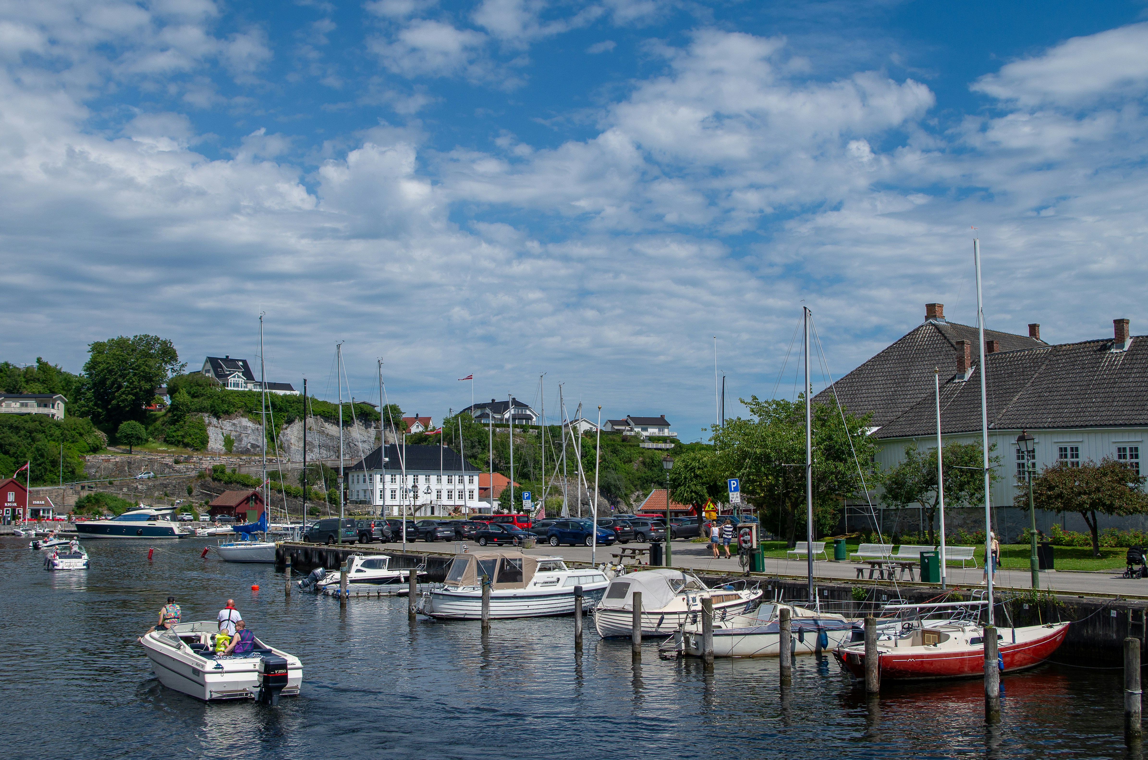 Harbor scene with docked boats and a coastal village backdrop under a partly cloudy sky.