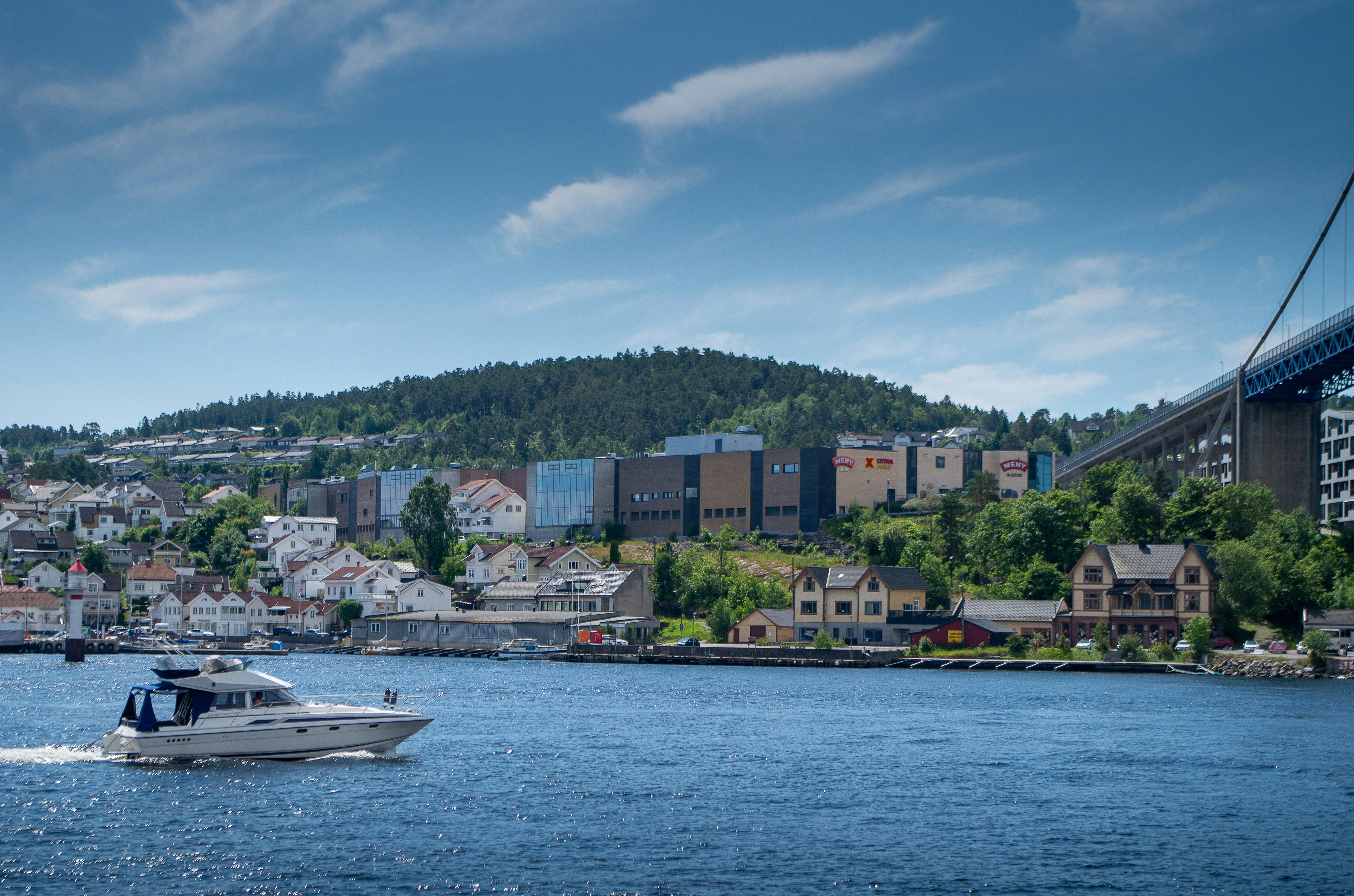 Bright harbor scene with a motorboat cruising past a waterfront town under a clear sky. Wide-frame composition emphasizes the river, buildings, and a distant hillside bridge.