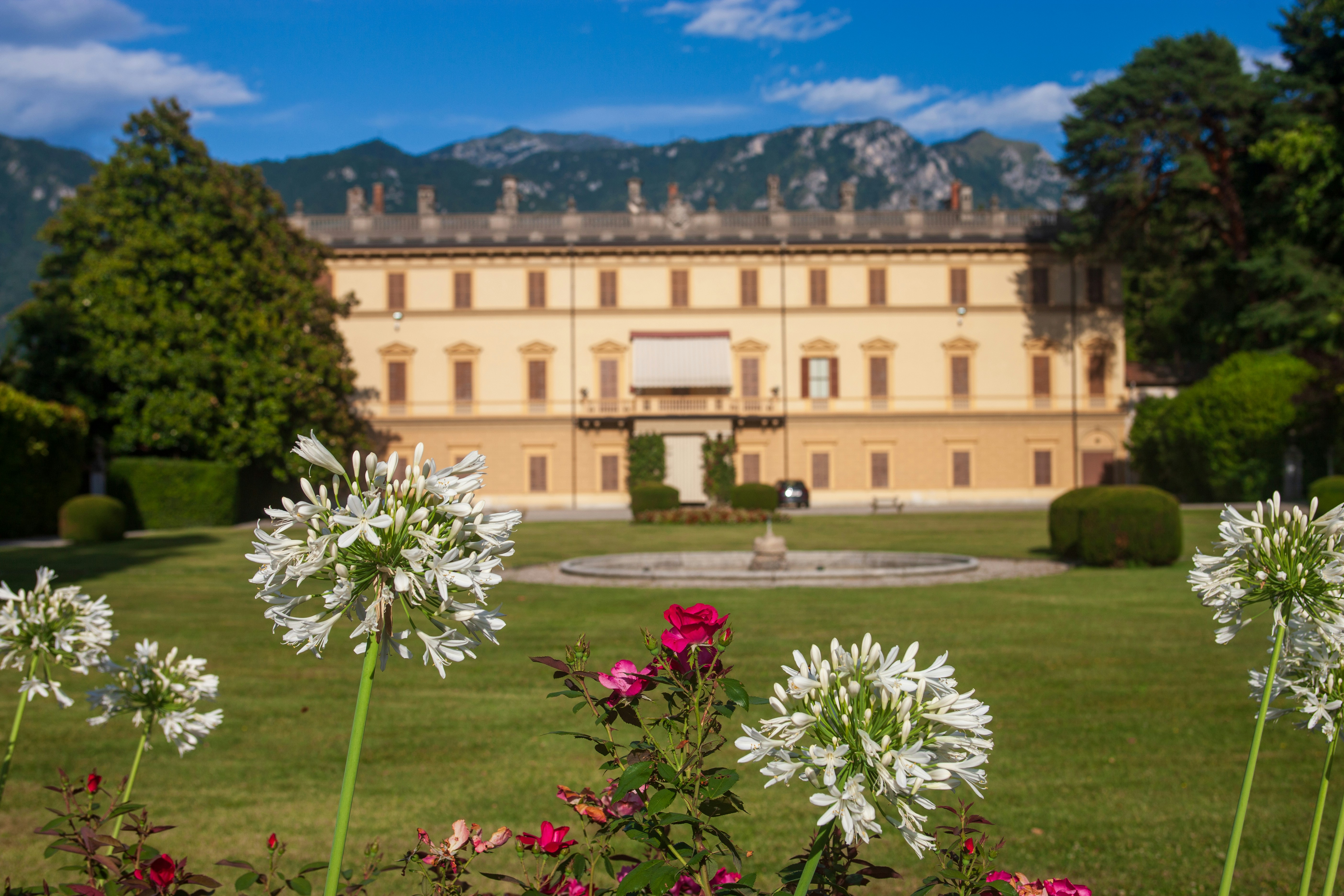 A grand chateau-like building sits behind a formal garden with white and pink flowers in the foreground, under a clear blue sky. The scene emphasizes architectural symmetry and manicured grounds.