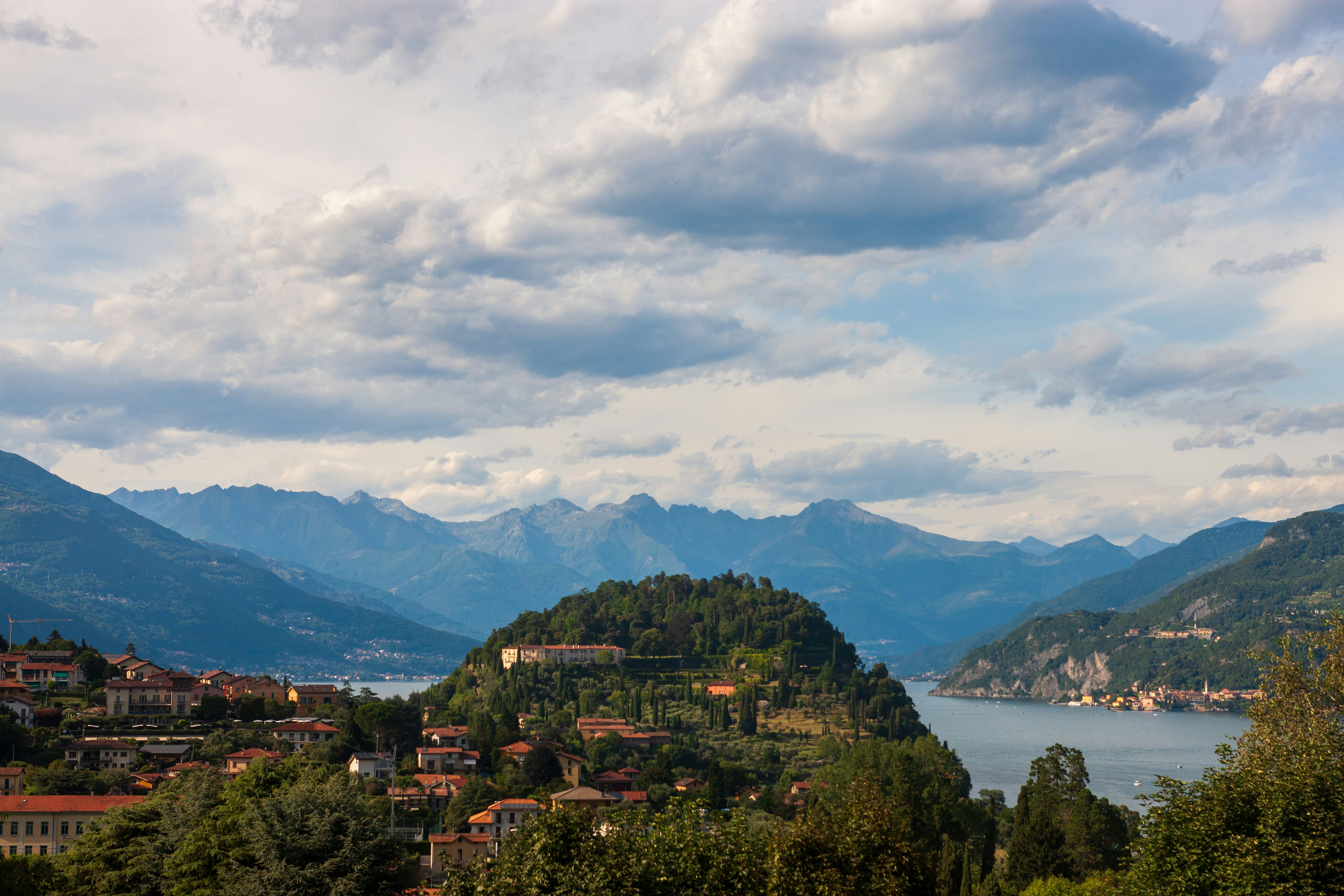 A tranquil hillside village perched above a lake, with layered mountains in the distance under a partly cloudy sky. The scene emphasizes the town’s red rooftops and verdant slopes against a vast, blue backdrop.